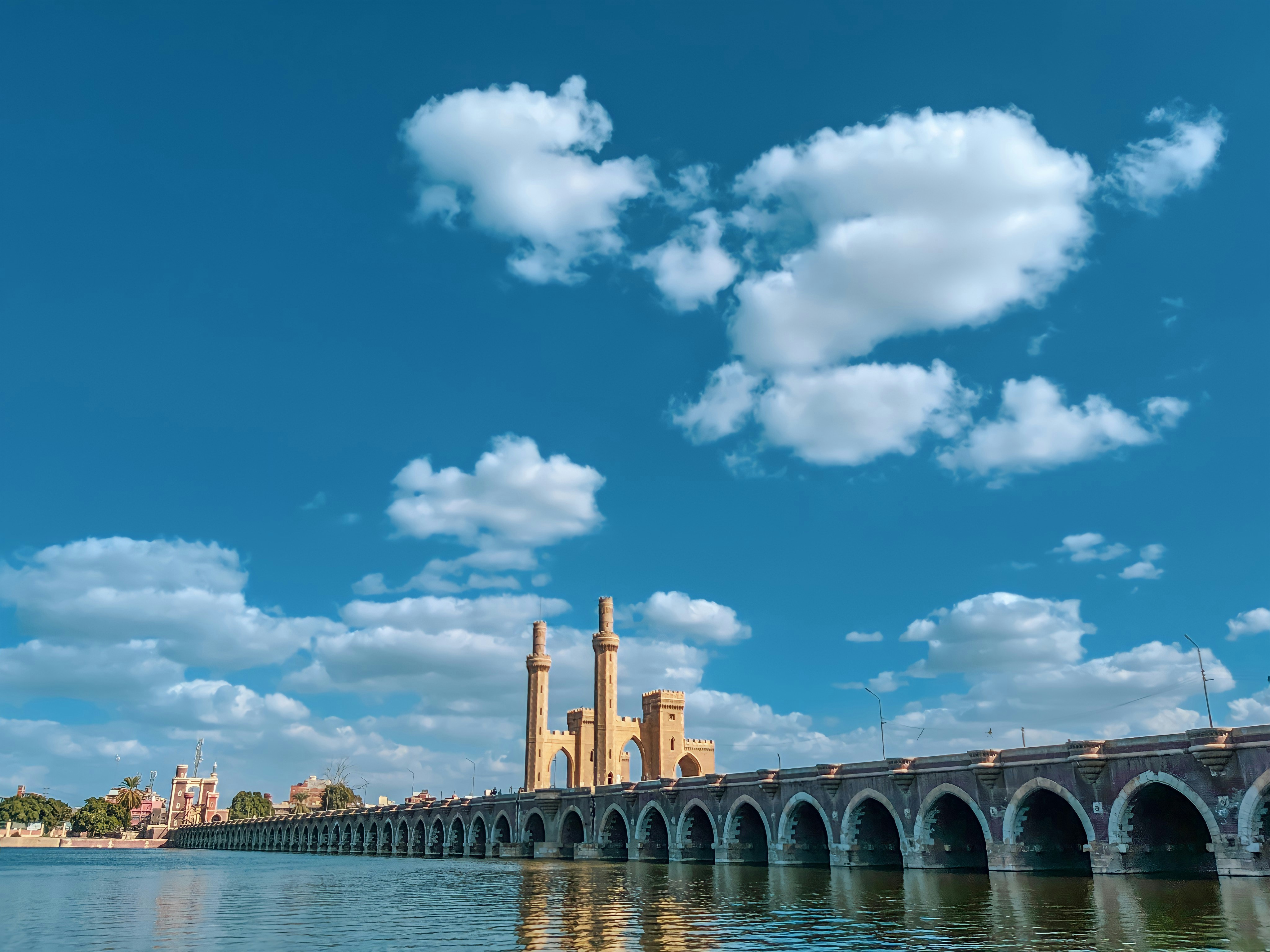 Historic stone bridge with arches stretching over water, flanked by distant towers under a bright blue sky with scattered clouds.
