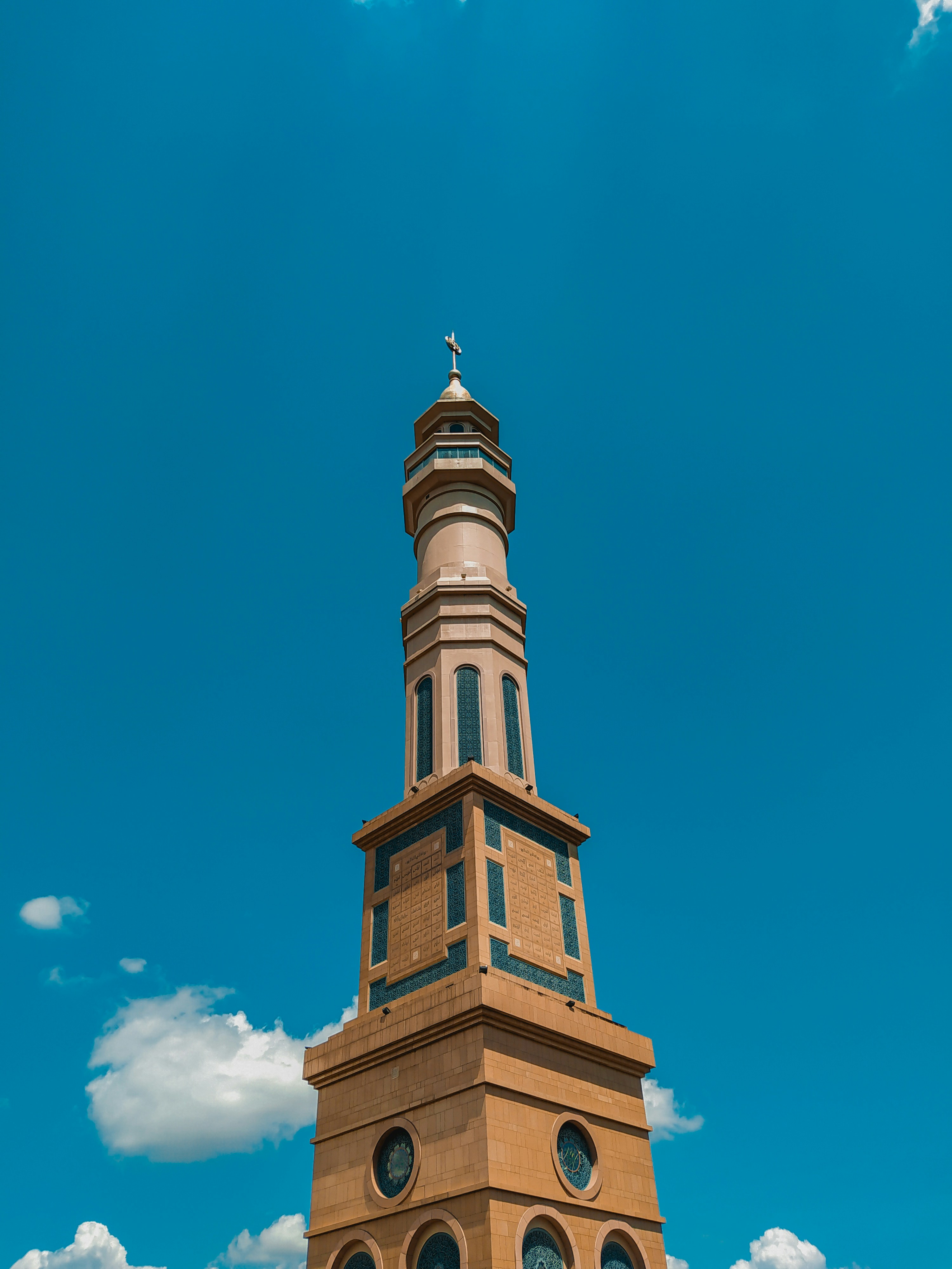 Tall architectural tower with a cross at the top, set against a bright blue sky dotted with clouds.