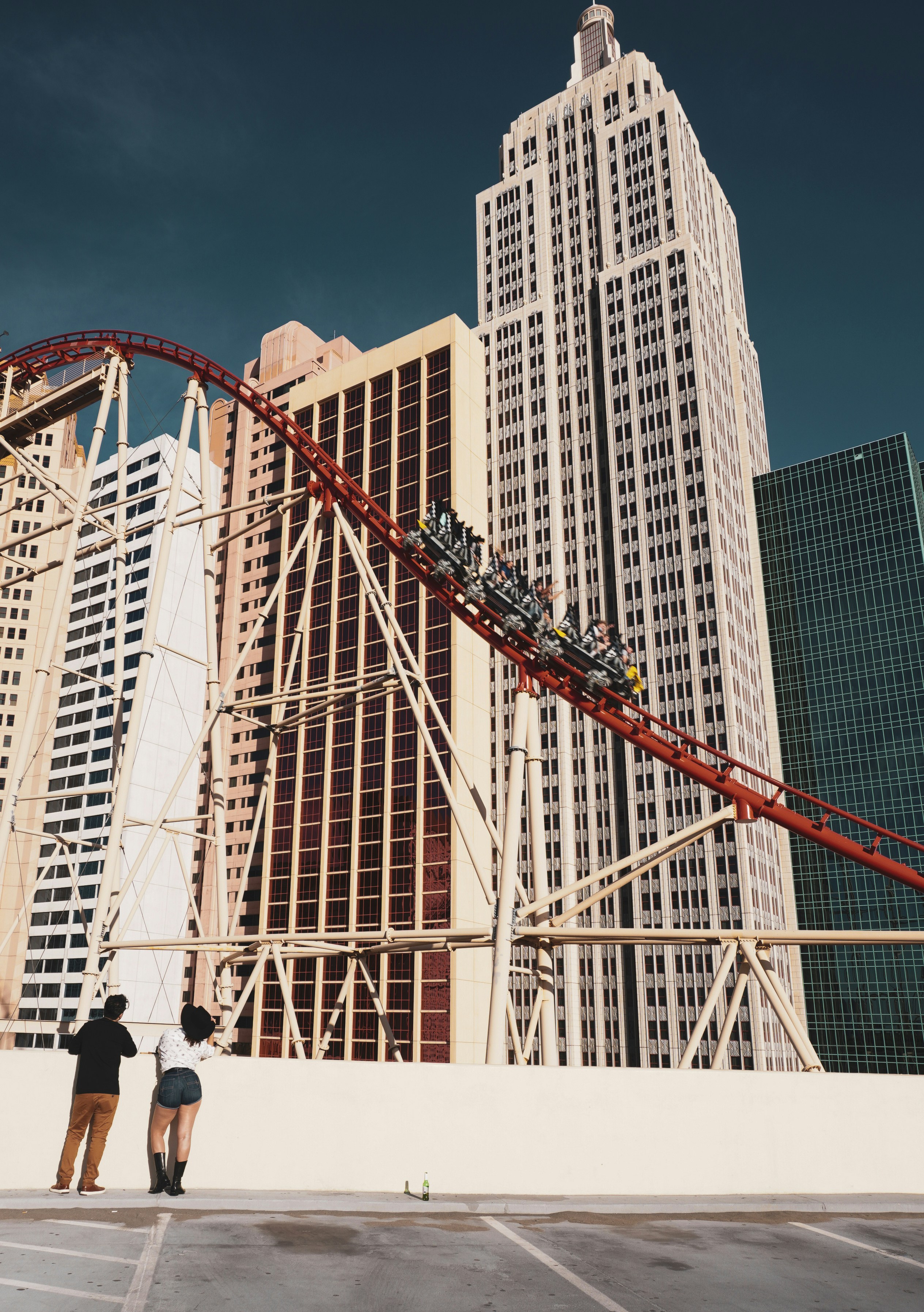 A roller coaster twists dramatically against a backdrop of towering skyscrapers, with two observers enjoying the view. The scene captures the excitement of urban amusement.