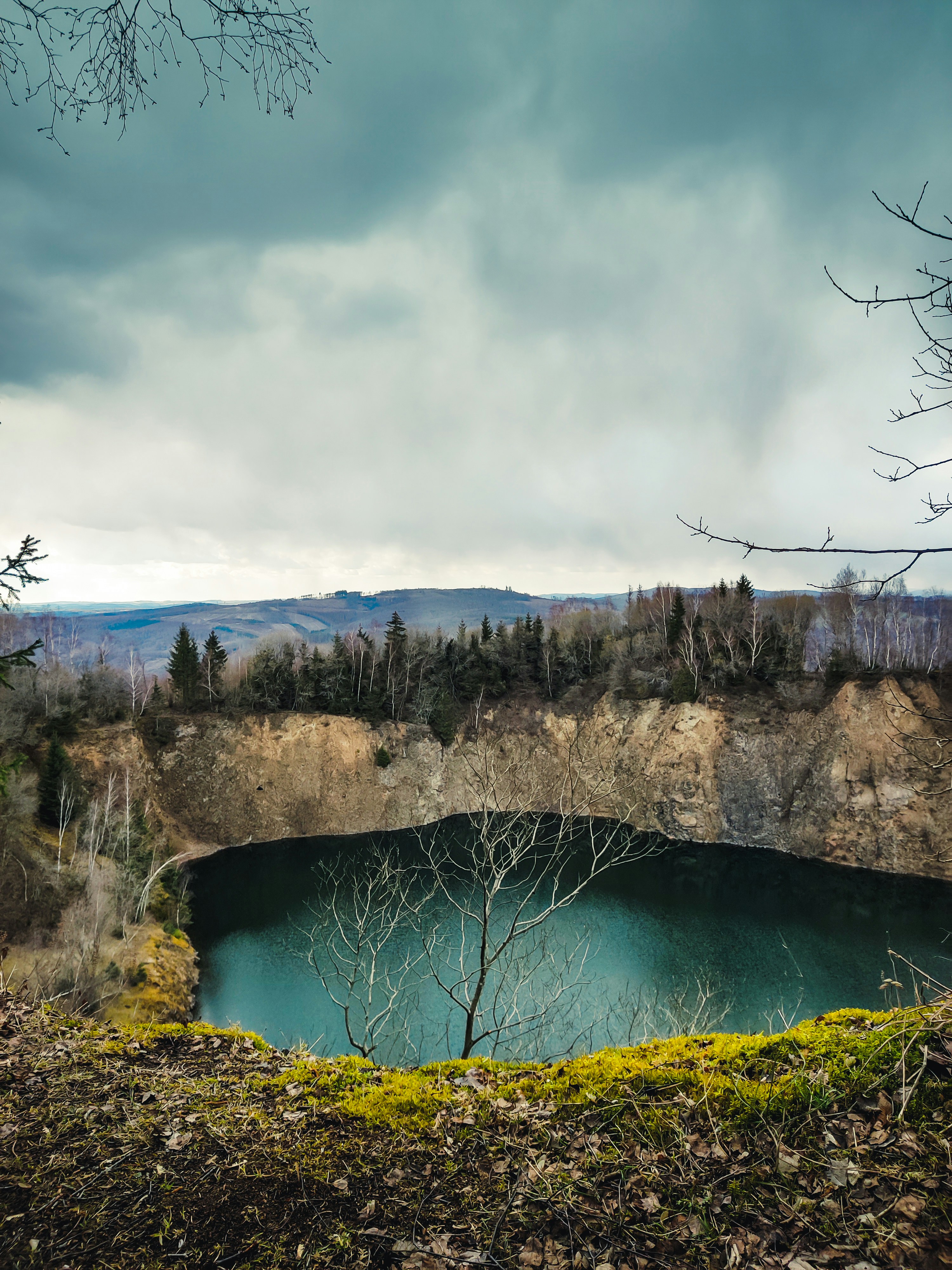 A tranquil quarry lake surrounded by bare trees and mossy ground under a dramatic, cloudy sky.