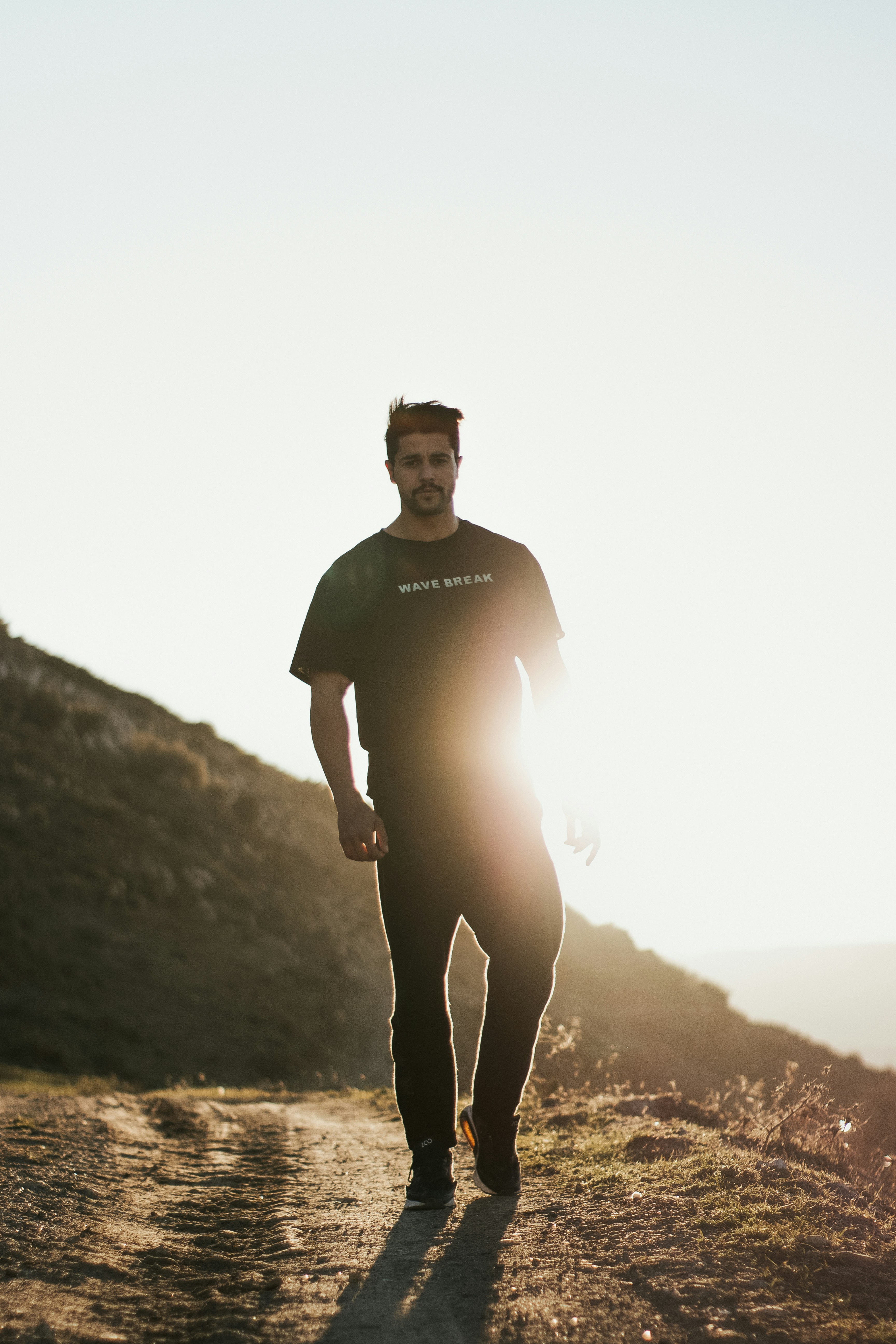 Silhouetted figure walking along a sunlit path, framed by a backdrop of rolling hills. The warm glow of the setting sun creates a striking contrast.