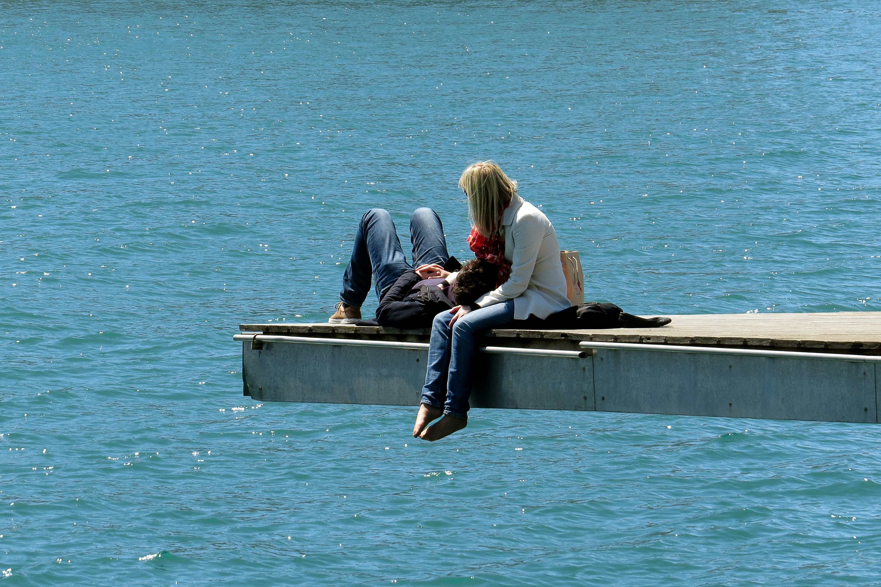 Three people relaxing on a concrete pier extending over a calm body of water under bright daylight.