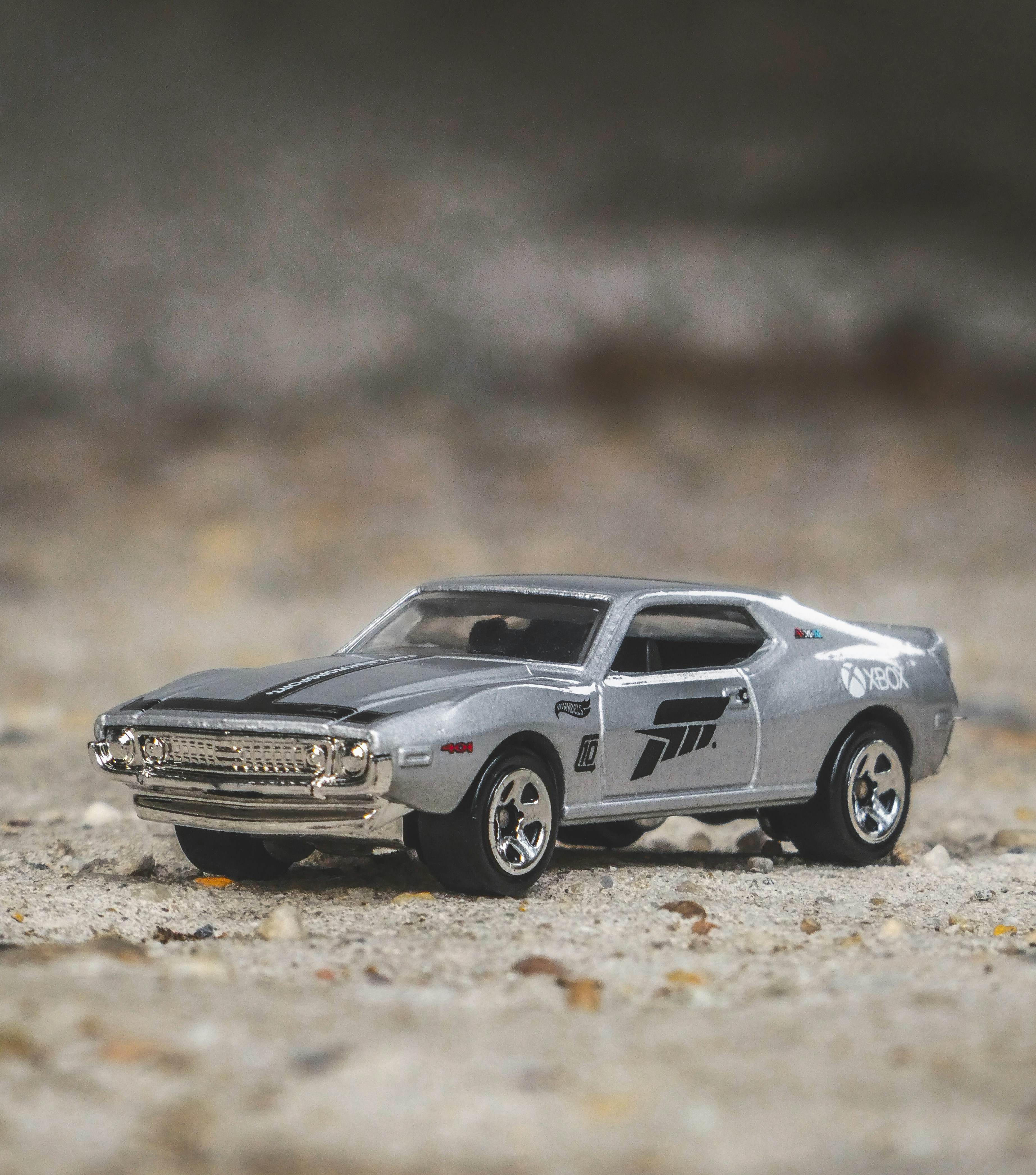 Detailed silver toy car with a sleek design, featuring racing stripes and logos, positioned on a textured surface.