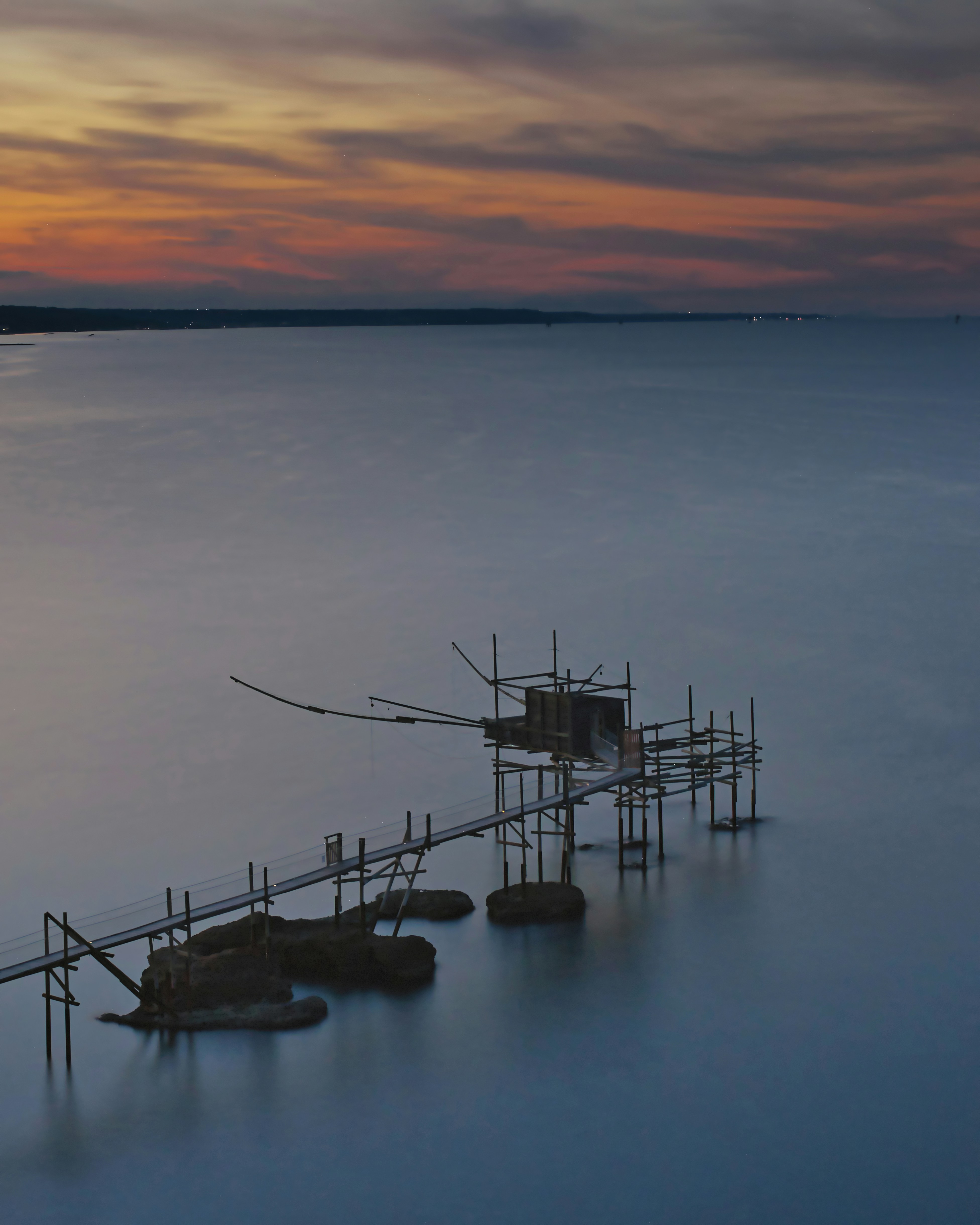 A wooden fishing structure extends into calm waters under a twilight sky, blending hues of orange and blue as day transitions to night.