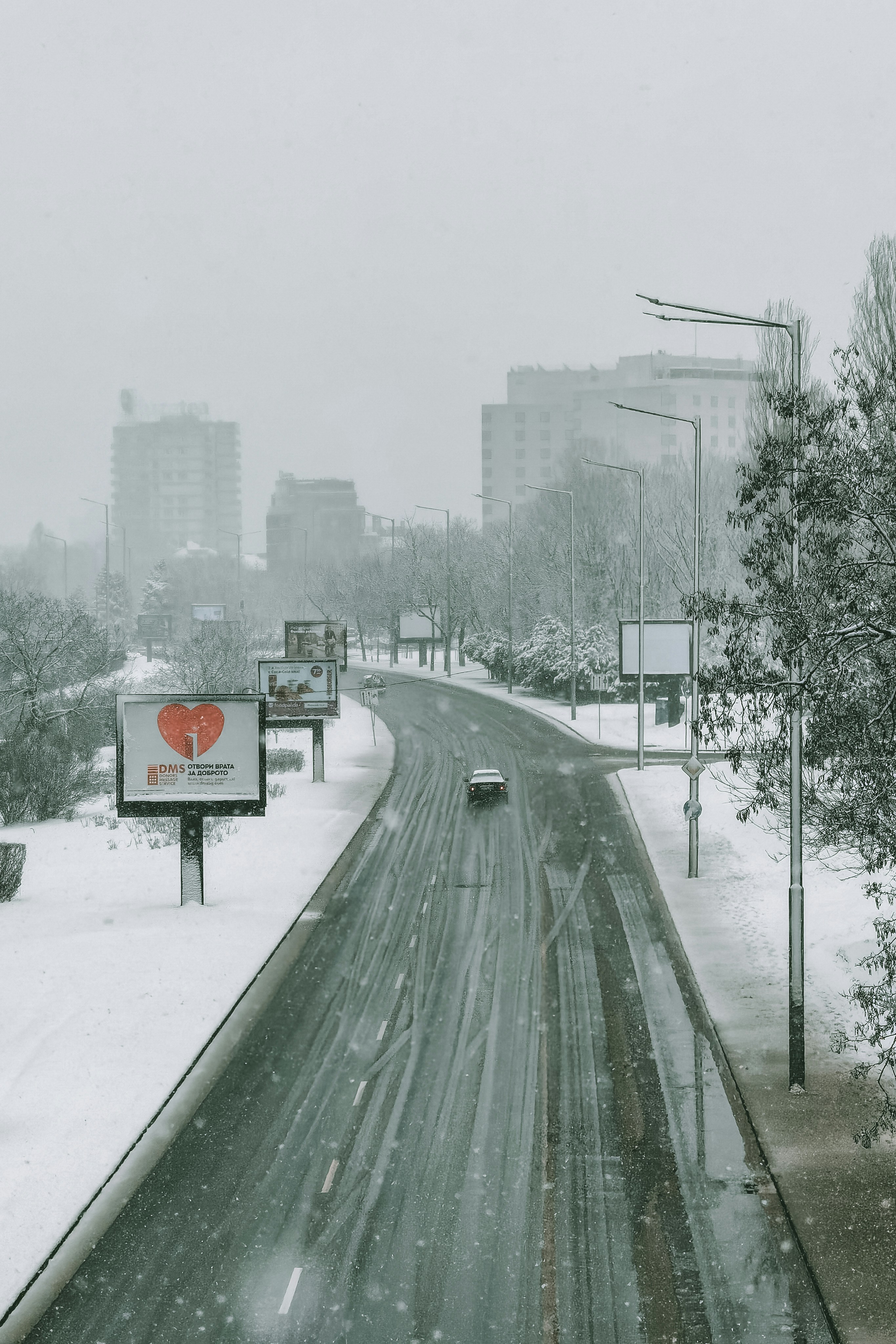 Snow falls over a quiet city boulevard as a lone car travels along a curved, wet road toward distant buildings.