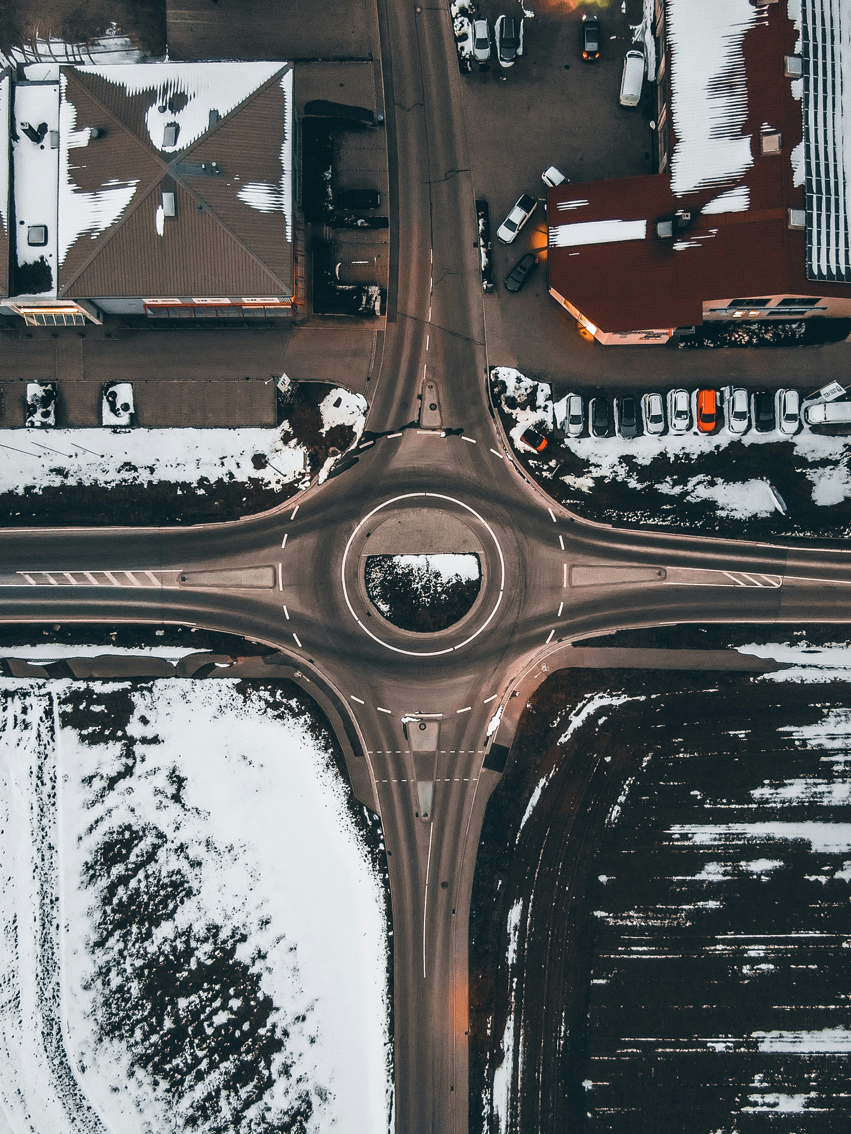 Aerial view of a roundabout surrounded by snowy fields and parked vehicles, highlighting the intersection's design and the contrast between urban and rural elements.
