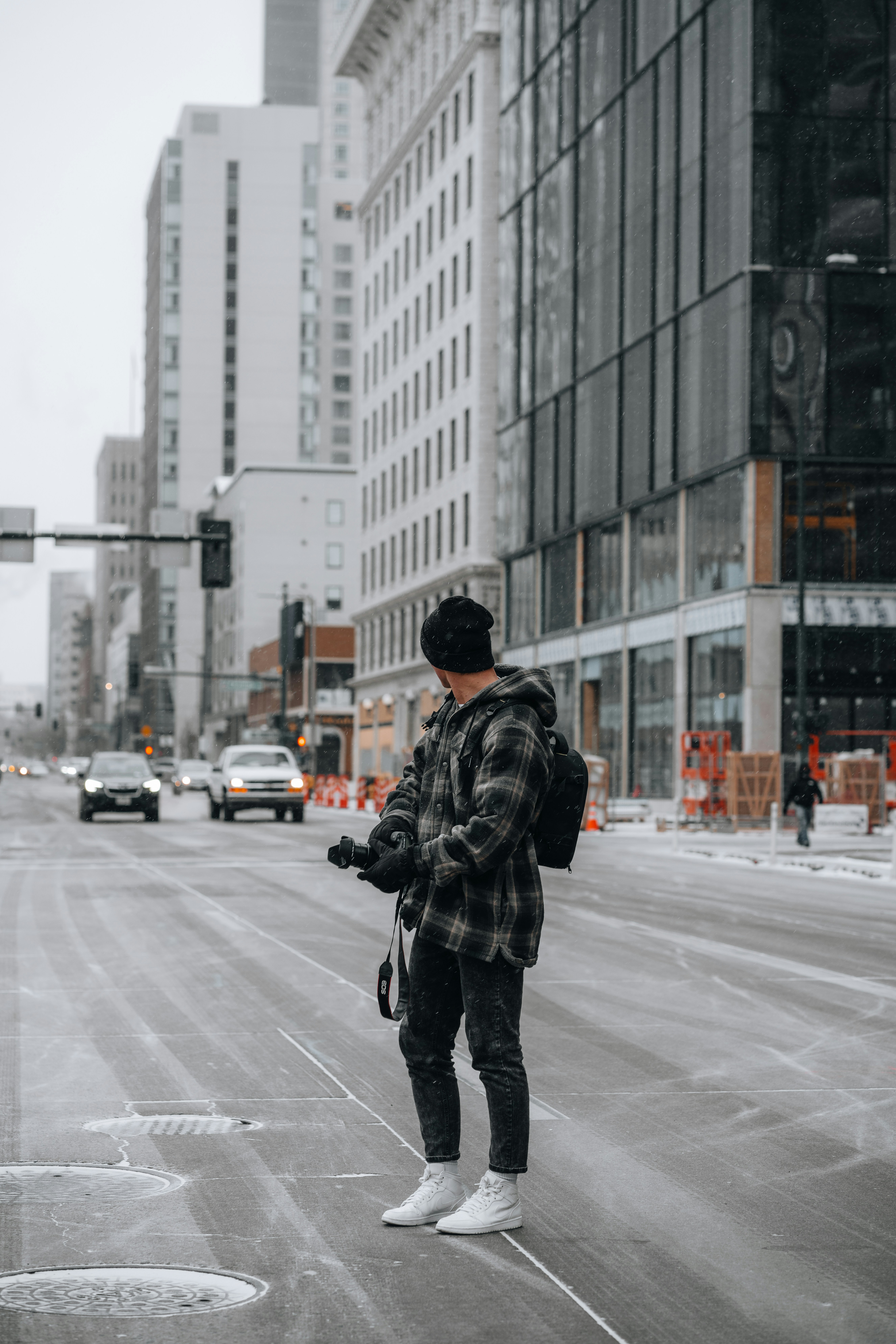 A photographer stands in a snowy urban street, capturing the essence of city life amidst falling snowflakes. The scene is framed by towering buildings and a muted color palette.