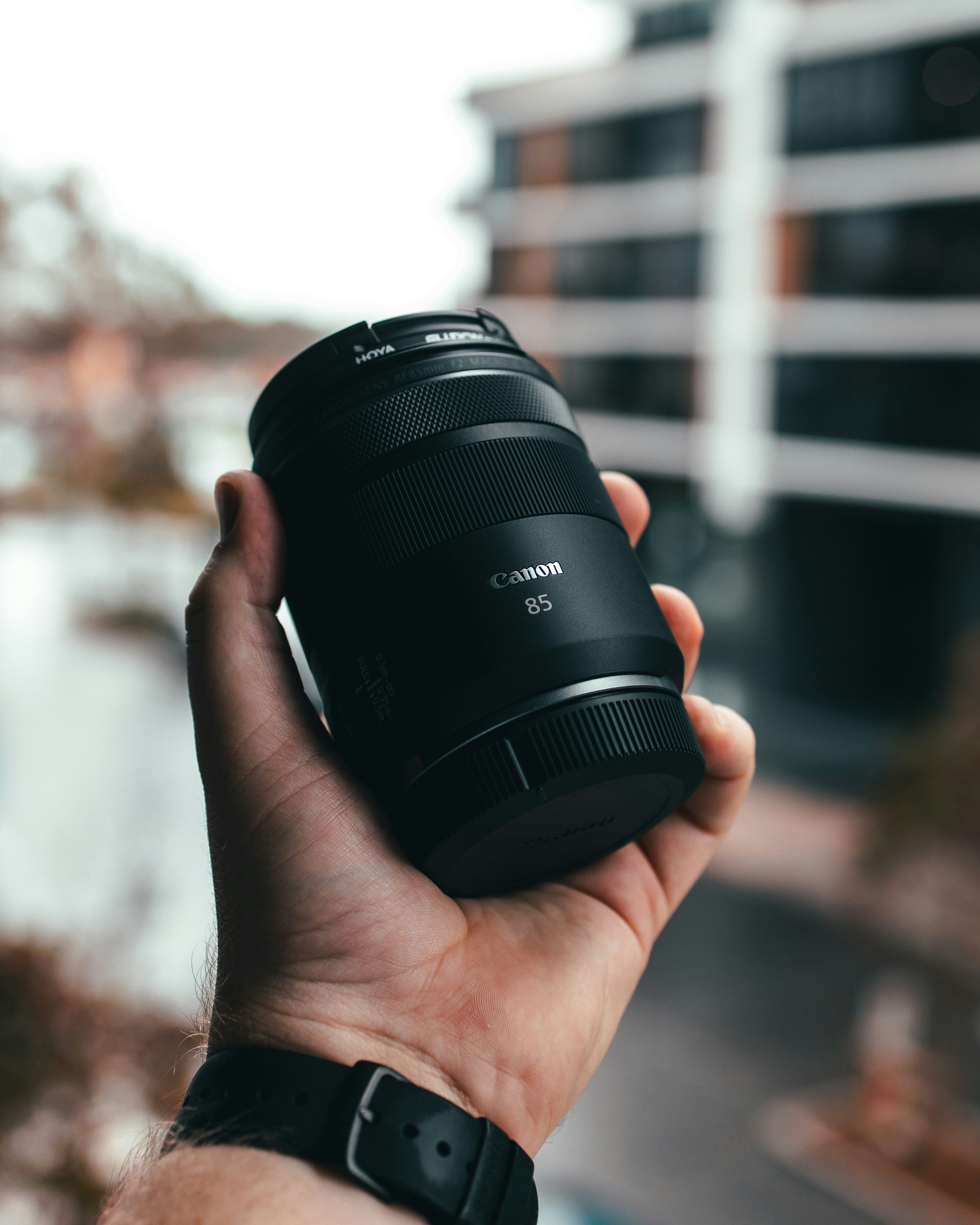 Hand holding a Canon 85mm lens against a blurred urban backdrop, showcasing the lens's design and build quality.