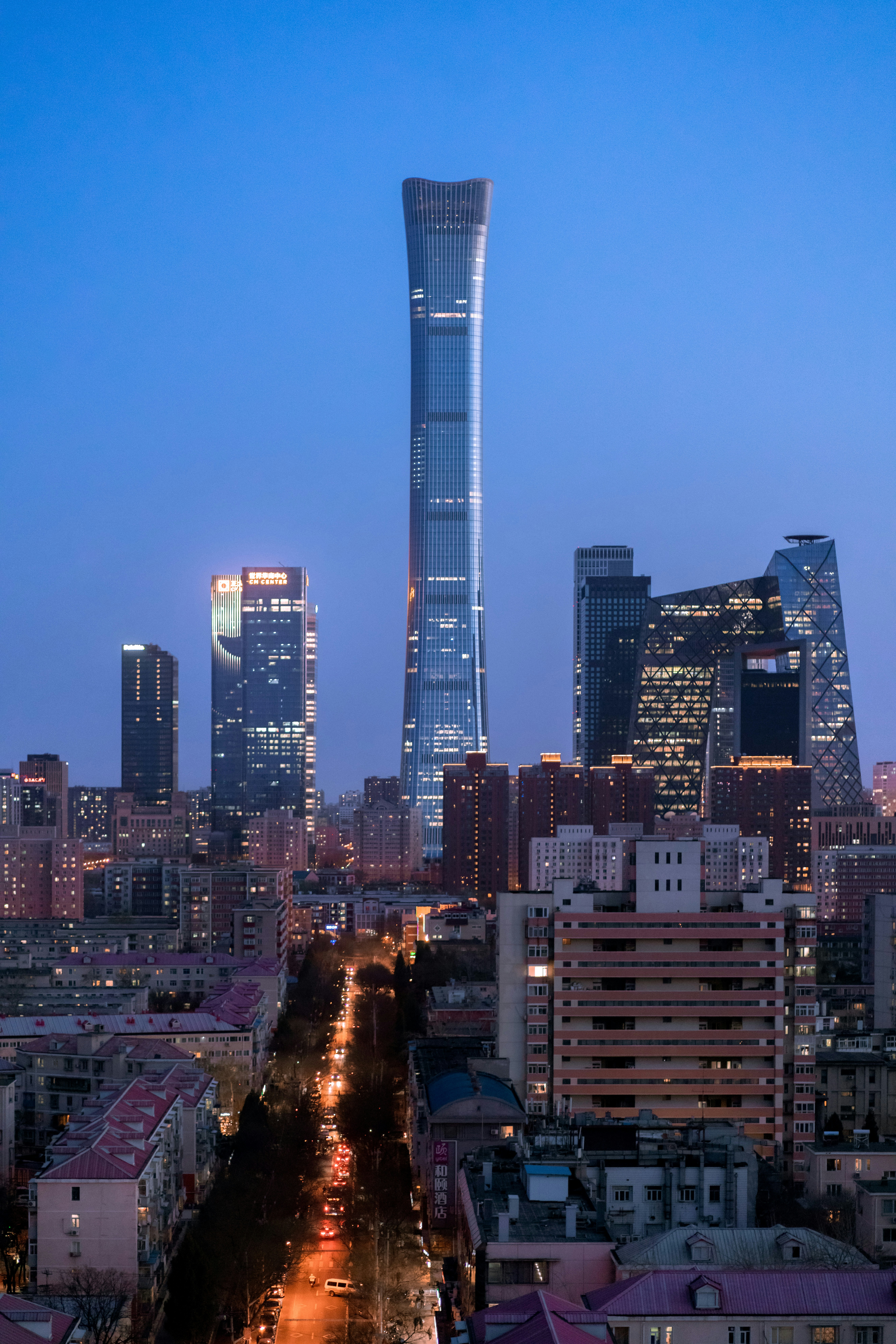 The skyline of Beijing at dusk, showcasing the iconic China Zun tower among other modern skyscrapers, with a busy street illuminated below.