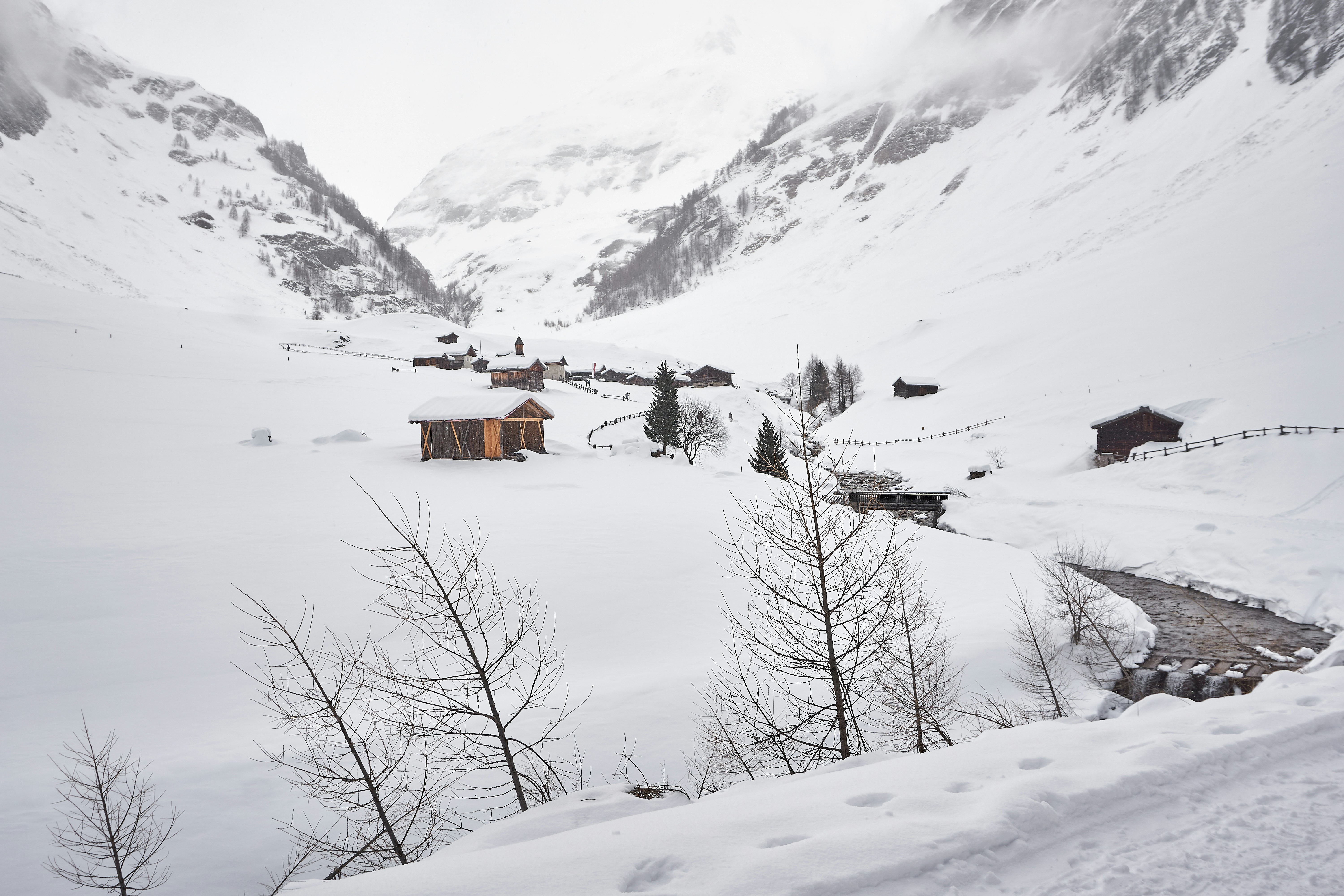 Snow-covered valley dotted with rustic wooden cabins and a winding stream, enveloped in a tranquil winter atmosphere.