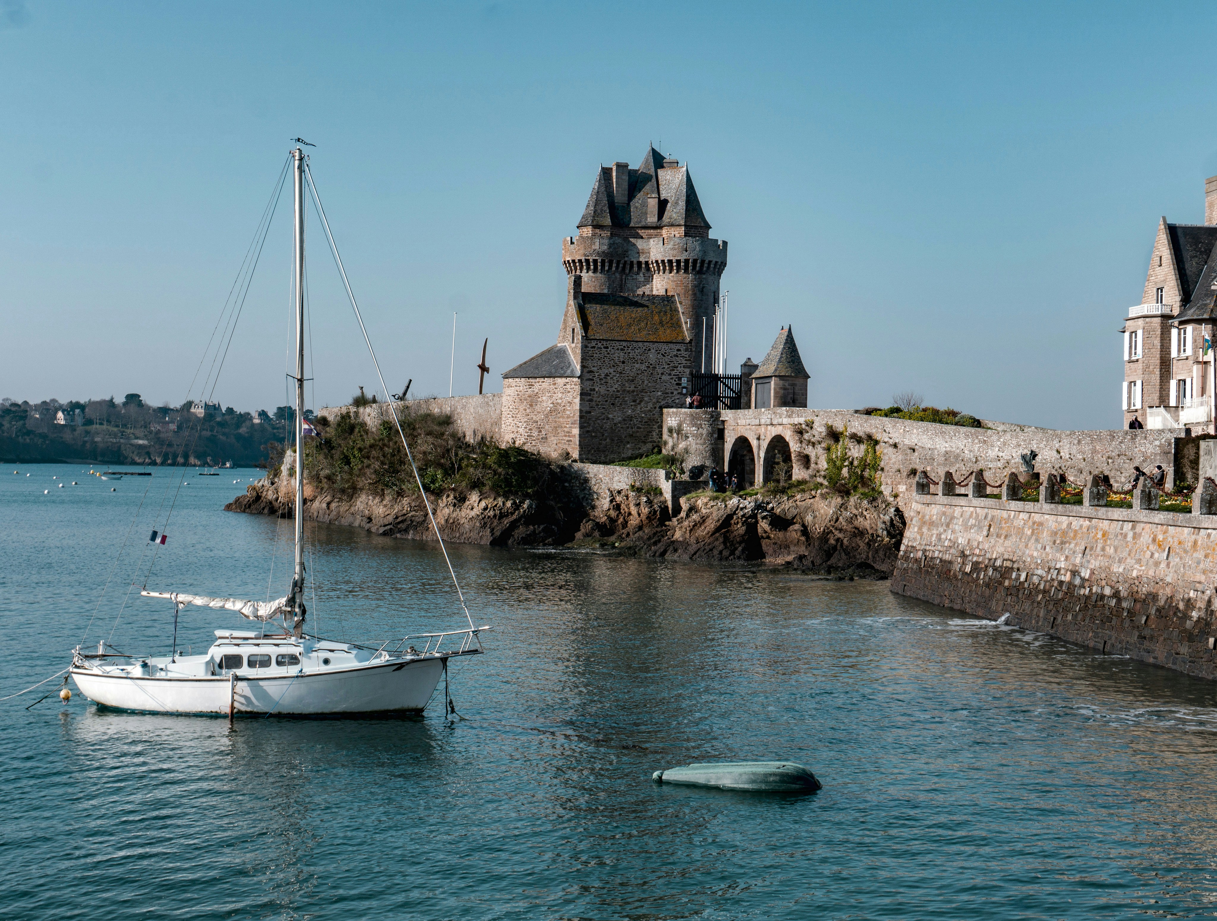 Sailboat anchored near a historic castle by the calm sea under a clear blue sky.