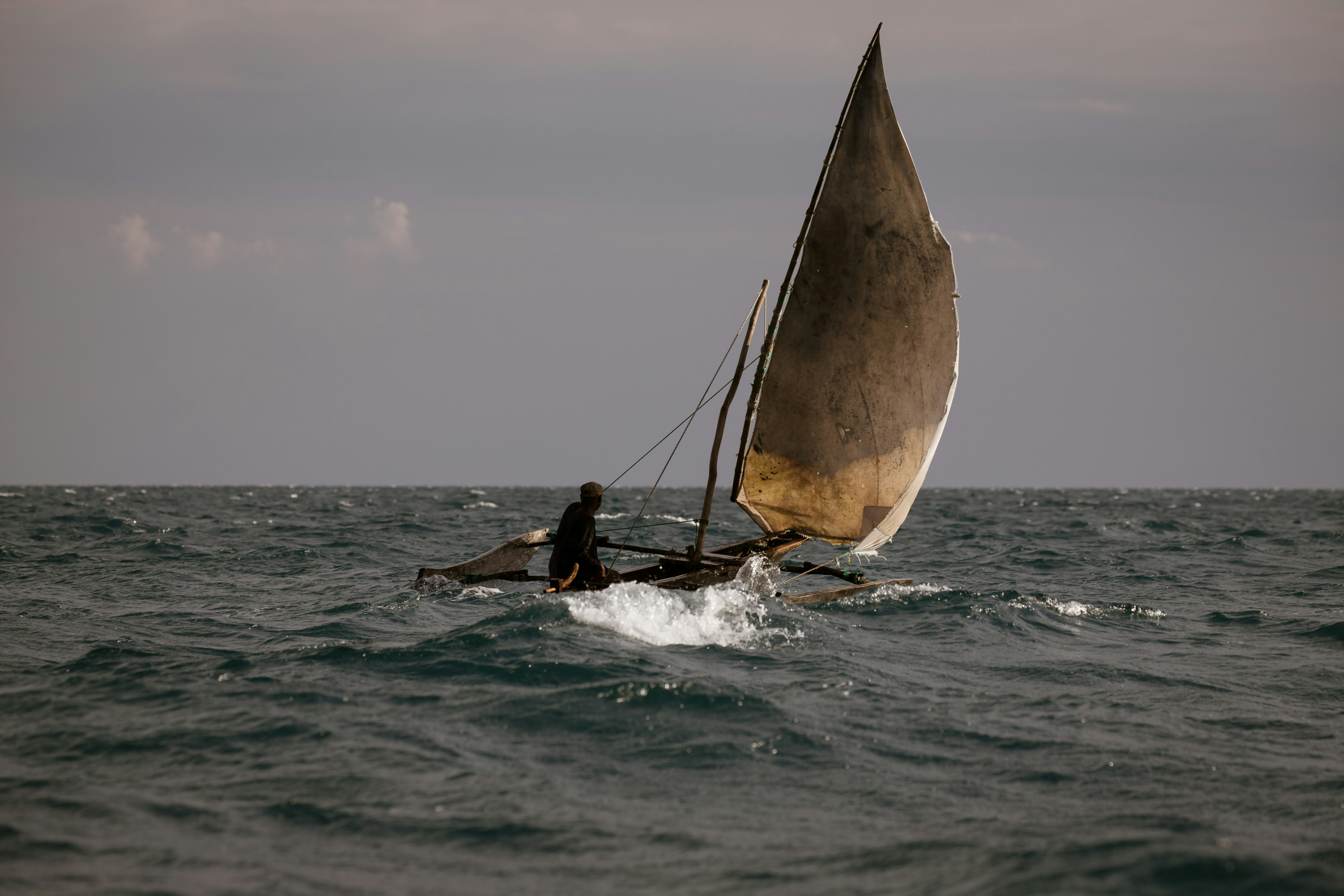 A lone sailor maneuvers a traditional boat through choppy waters, the wind filling the large sail against a moody sky.