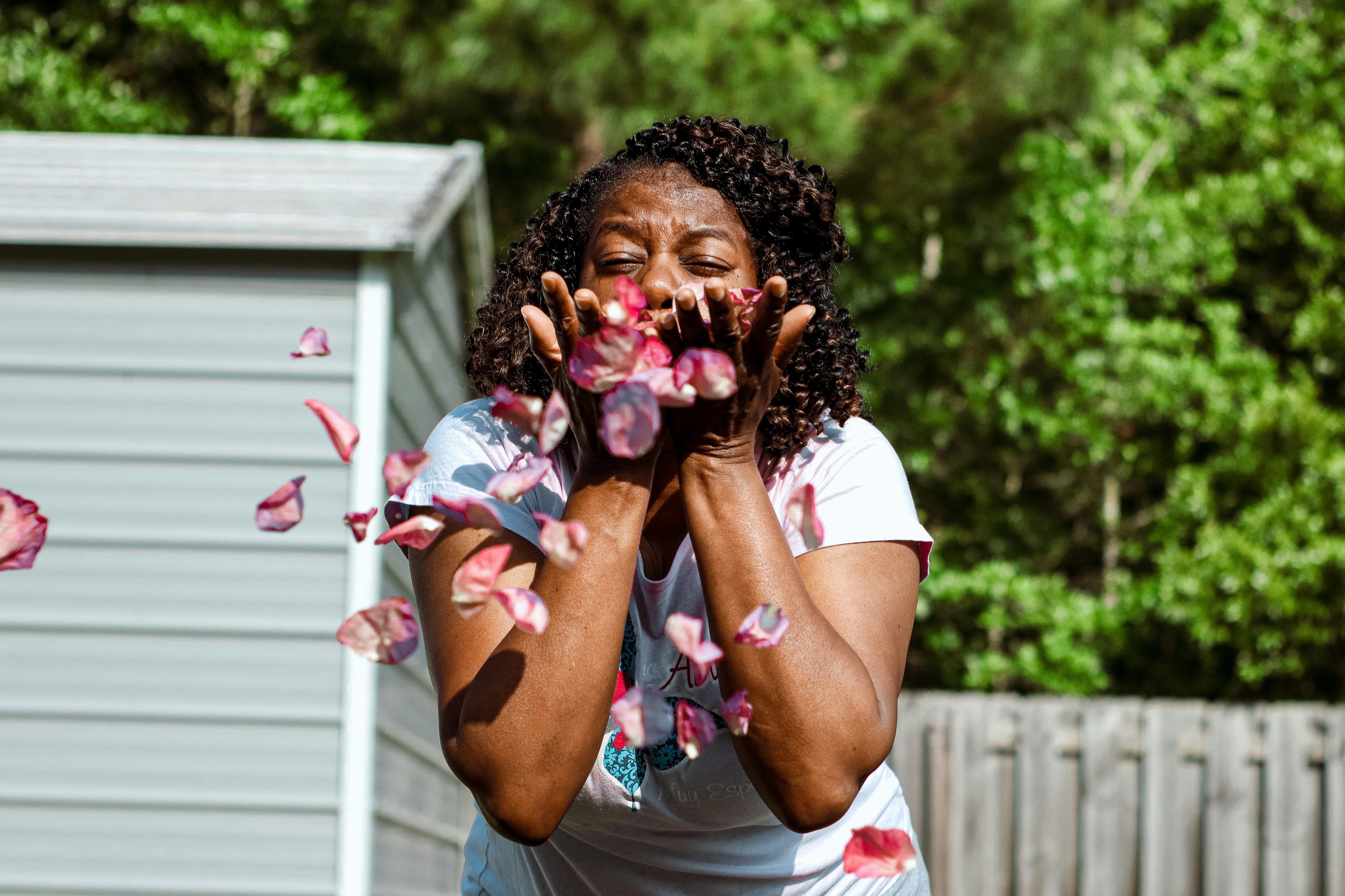Woman joyfully tossing rose petals into the air, surrounded by greenery. The scene captures a moment of happiness and connection with nature.
