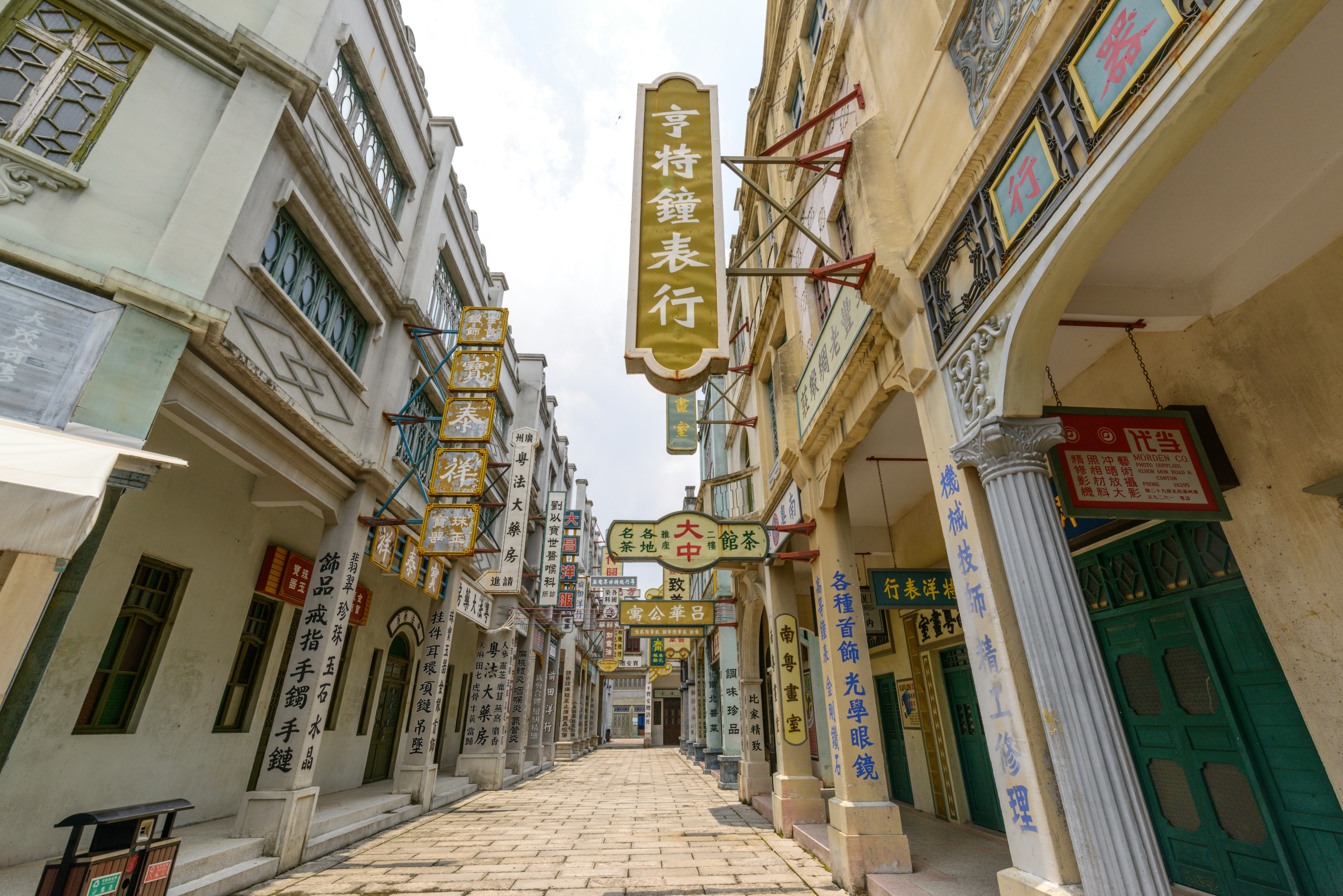 Narrow street in a historic district lined with ornate facades and traditional signage under a bright sky.