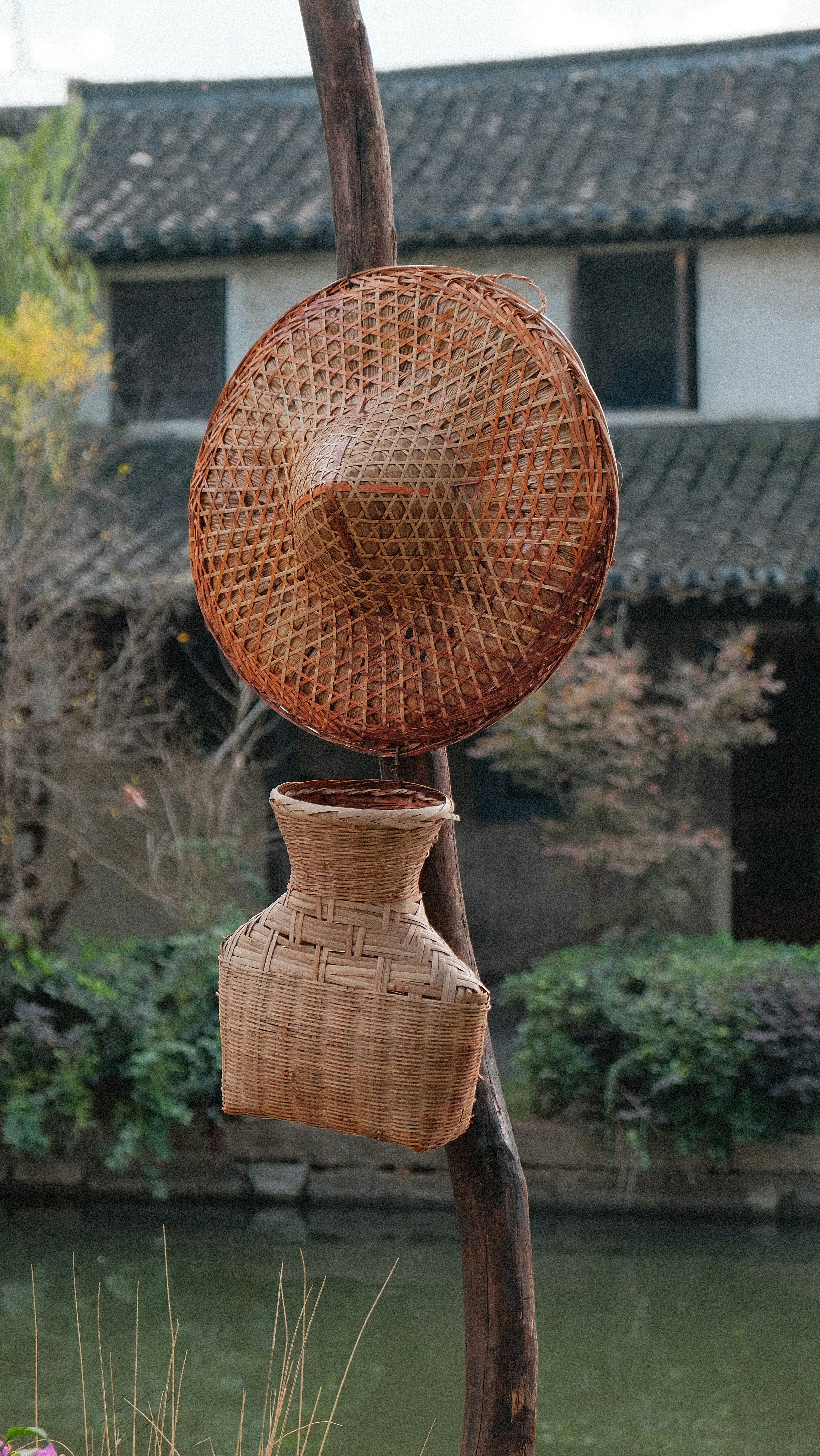 Intricately woven straw hat and basket hanging from a wooden post near a serene waterway, showcasing traditional craftsmanship.