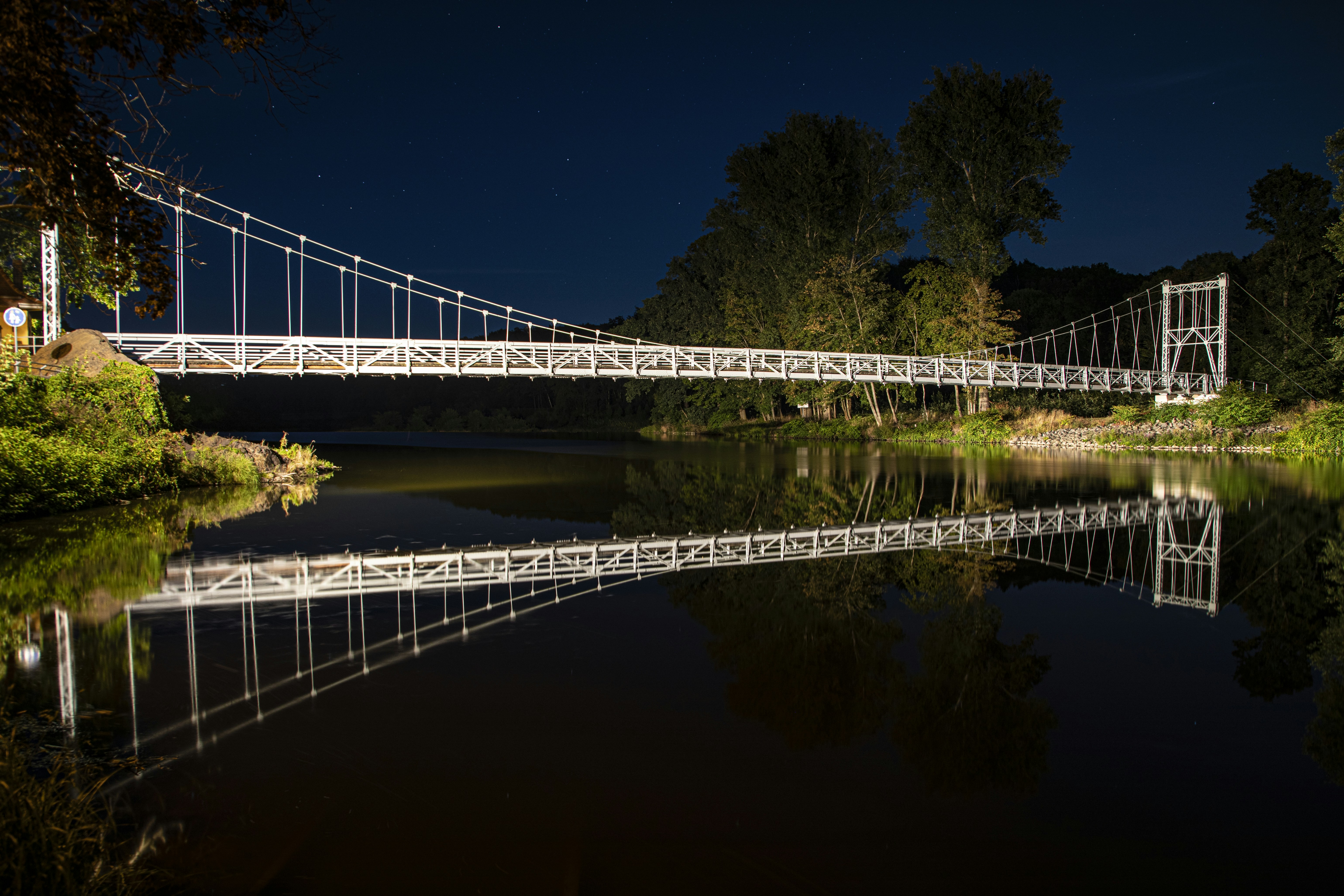 Illuminated suspension bridge reflected in calm waters under a starry sky.