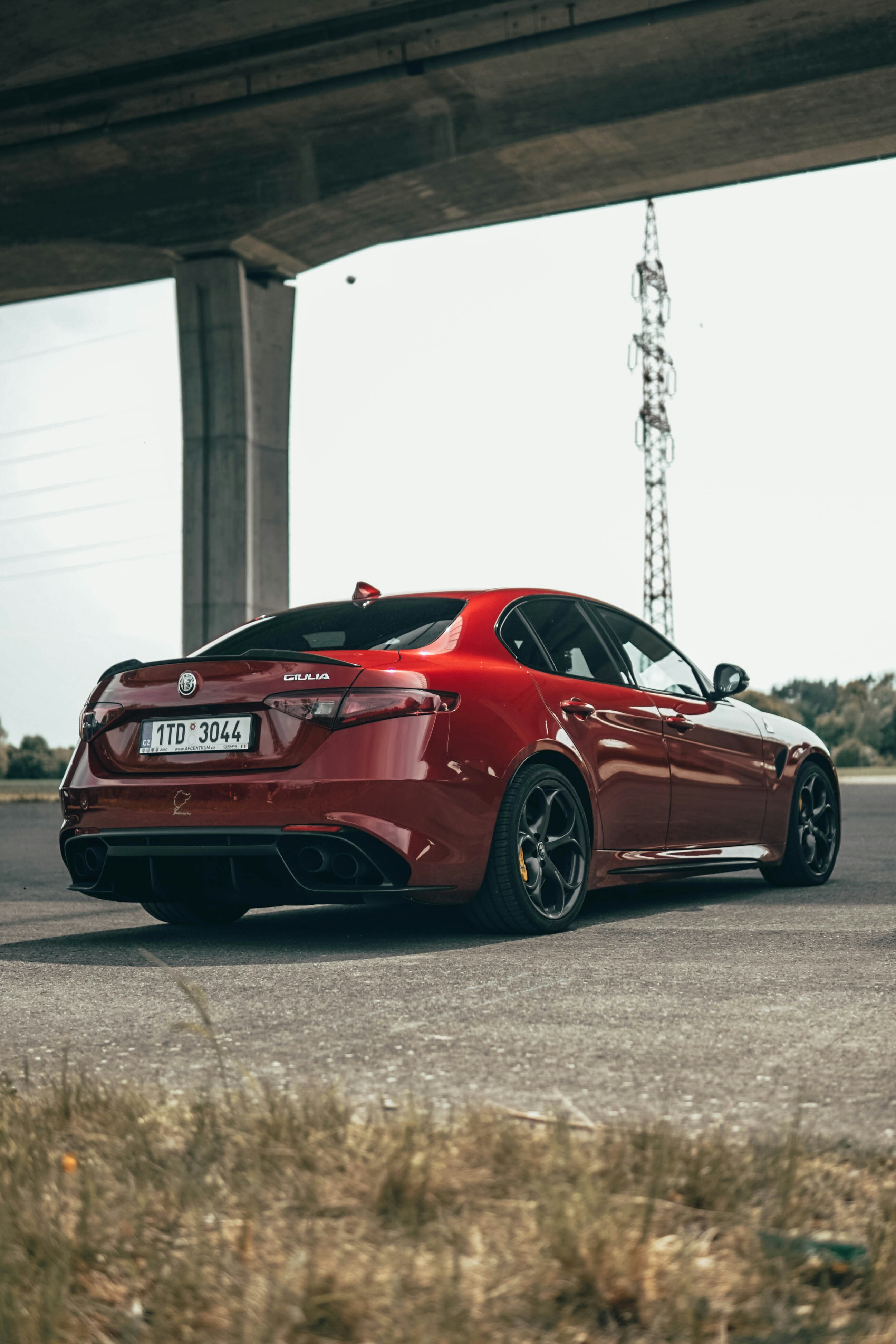 Red Alfa Romeo Giulia QV parked beneath a concrete overpass with a distant power line tower.
