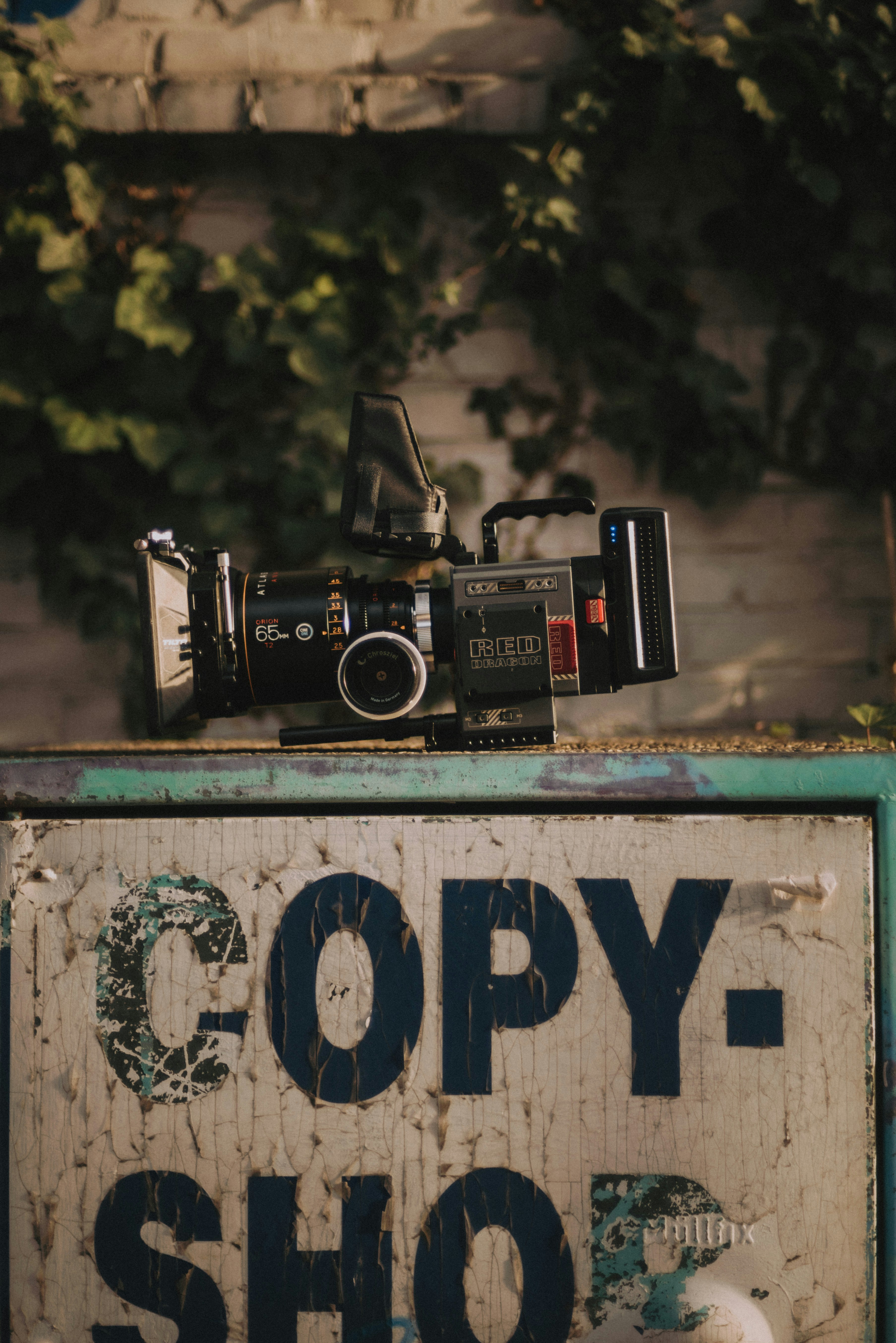 A RED camera positioned atop a weathered 'COPY SHOP' sign, framed by lush greenery in the background.