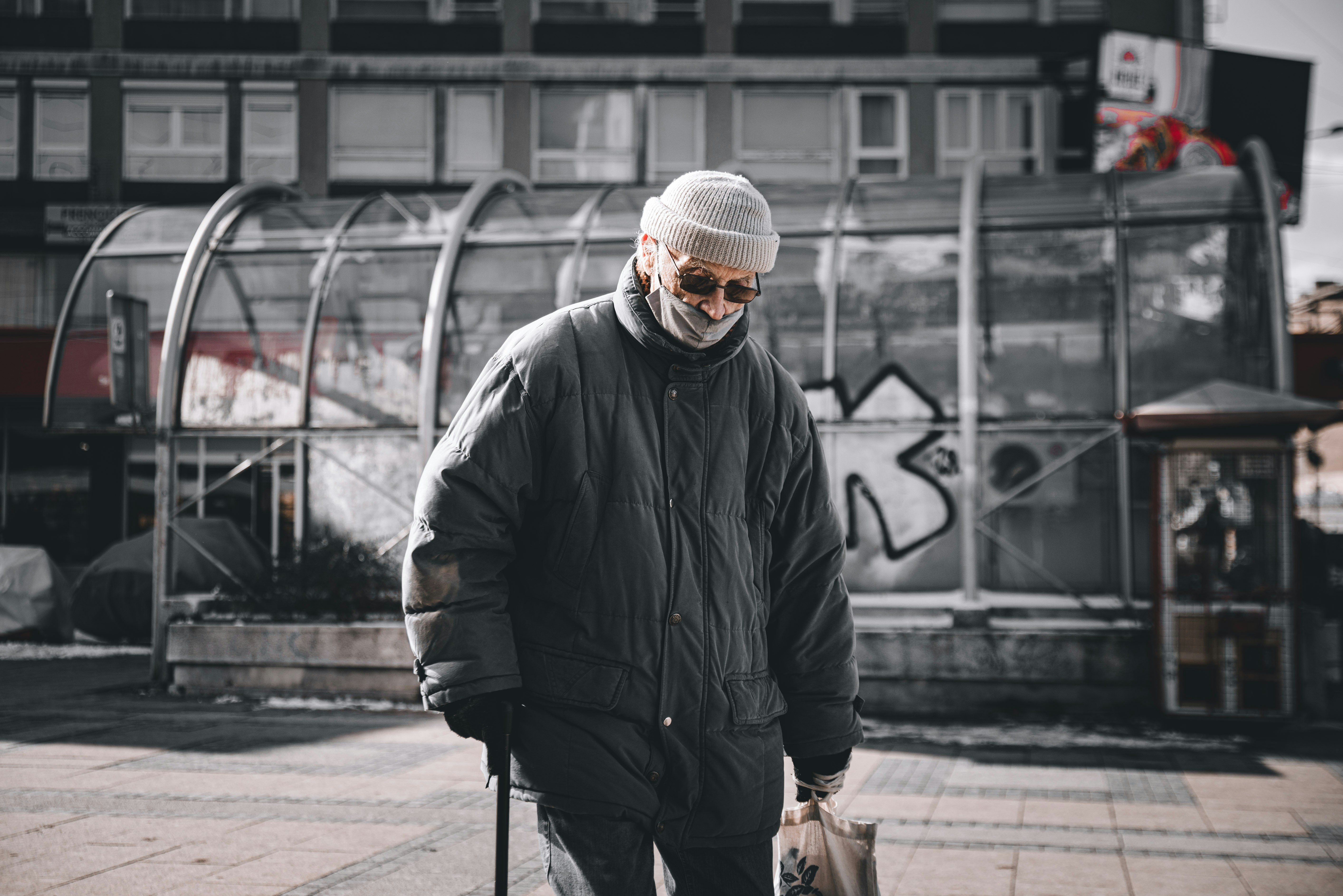 Elderly man in a winter coat walking with a cane in front of a graffiti-covered bus shelter.