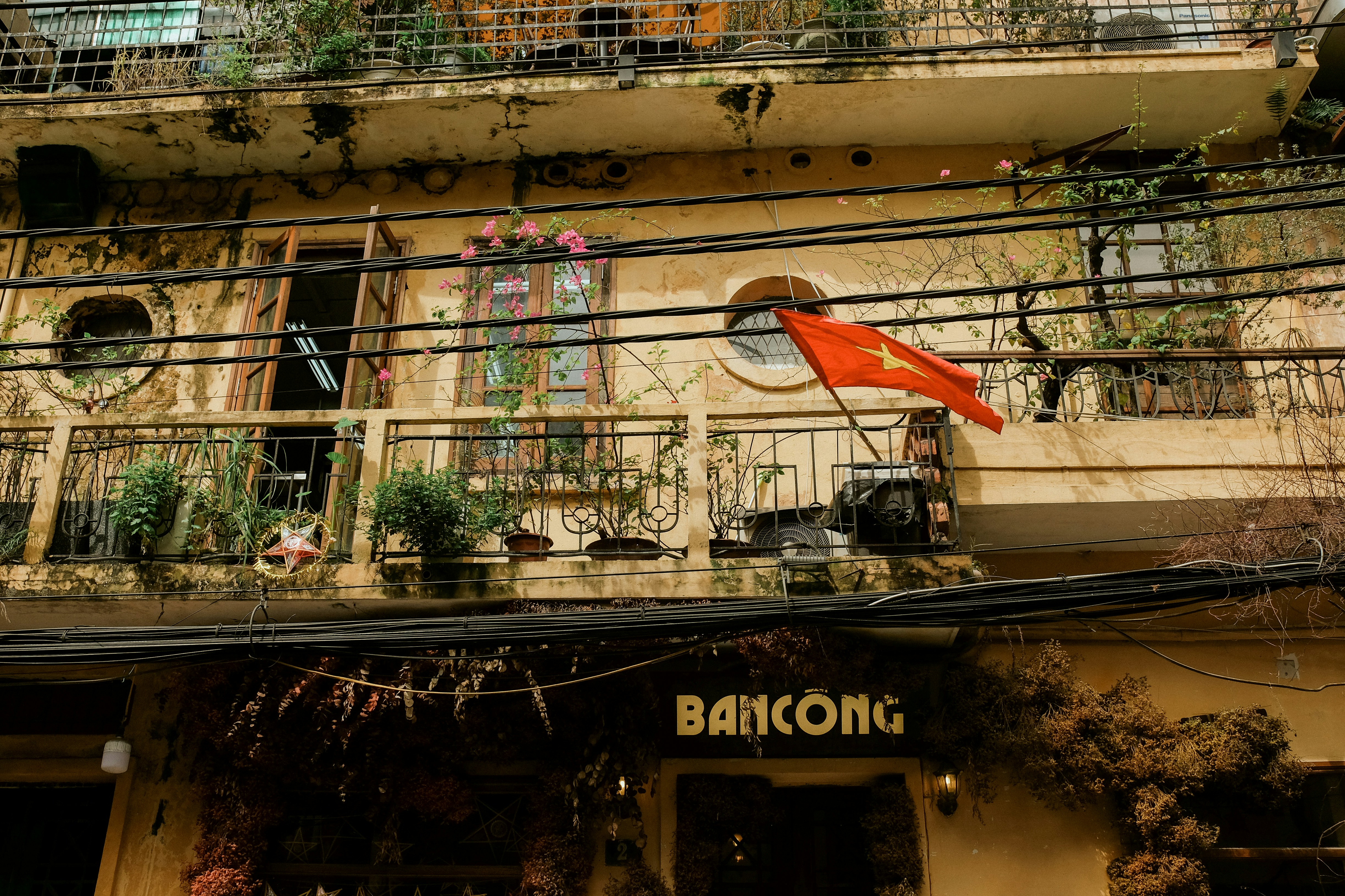 Balcony adorned with potted plants and a red flag on an aged Hanoi building.