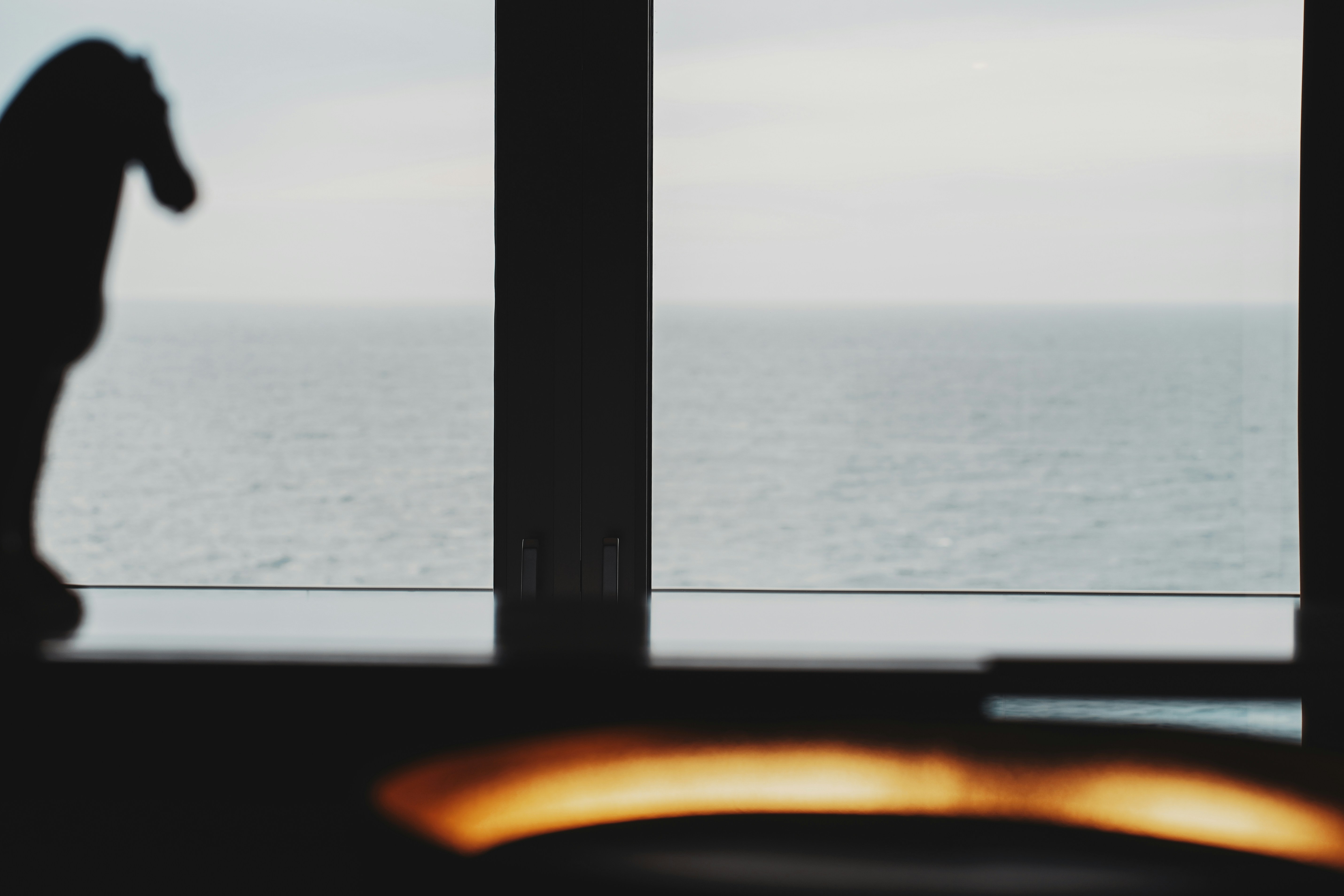 Silhouette of a horse figurine set against a vast ocean view through a window, capturing a moment of tranquility and reflection.