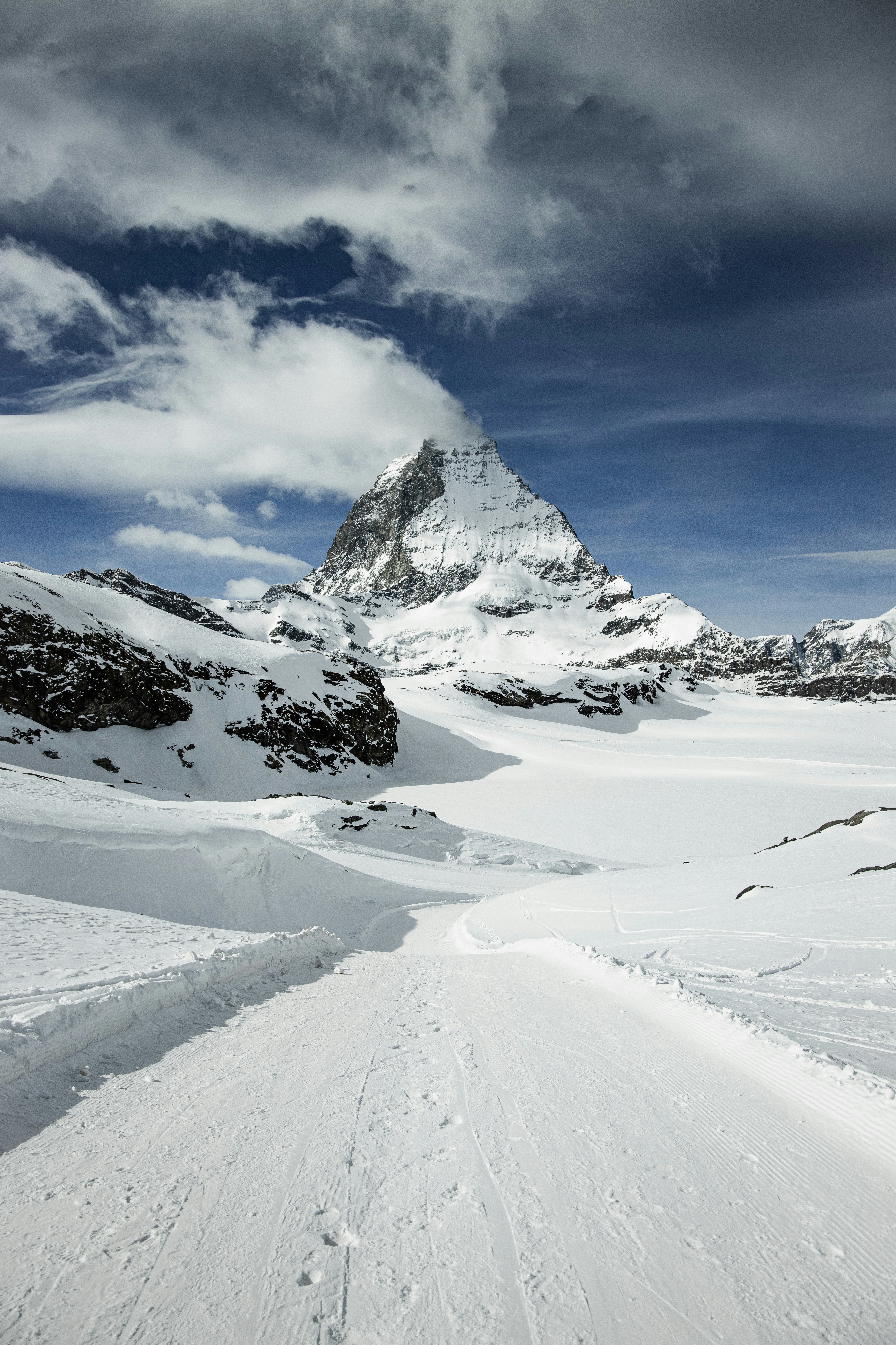 Snow-covered mountain peak rises dramatically against a backdrop of clouds and blue sky, with a winding path leading through the pristine snowfield.