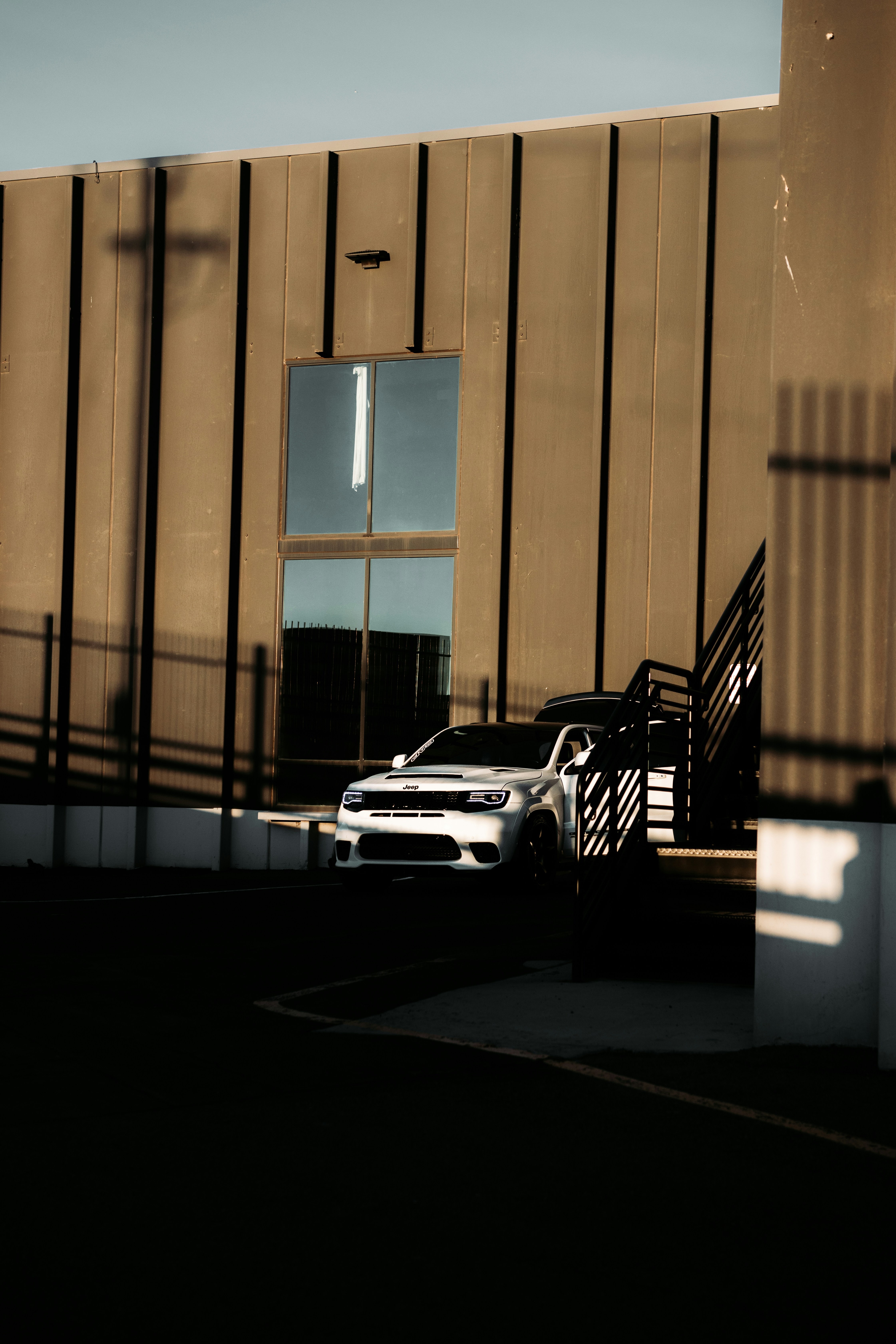 A sleek white vehicle parked beside a modern industrial building, casting sharp shadows on the pavement. The interplay of light and architecture creates a striking visual narrative.