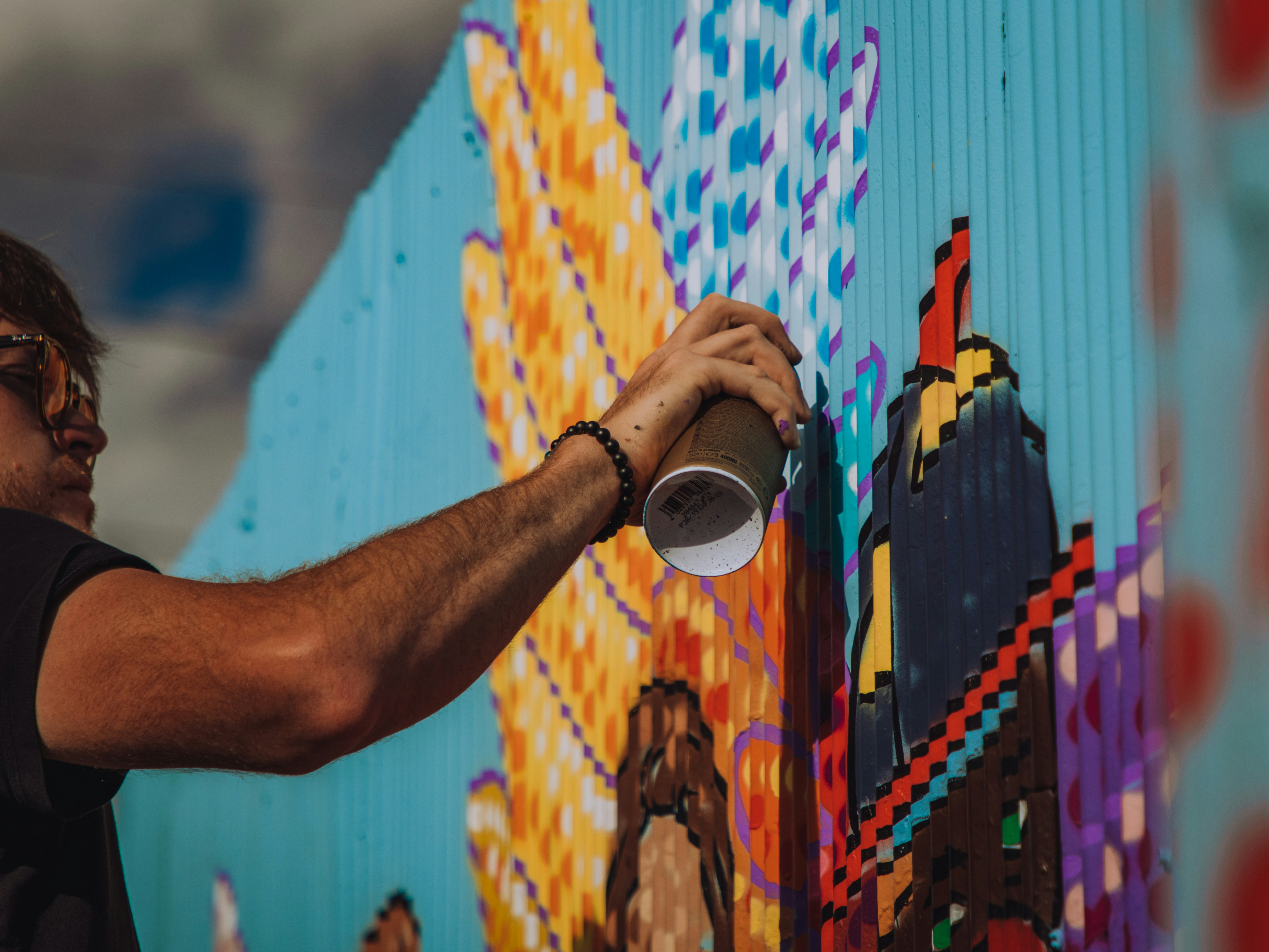 Street artist applying colorful spray paint to a mural on a corrugated metal surface.