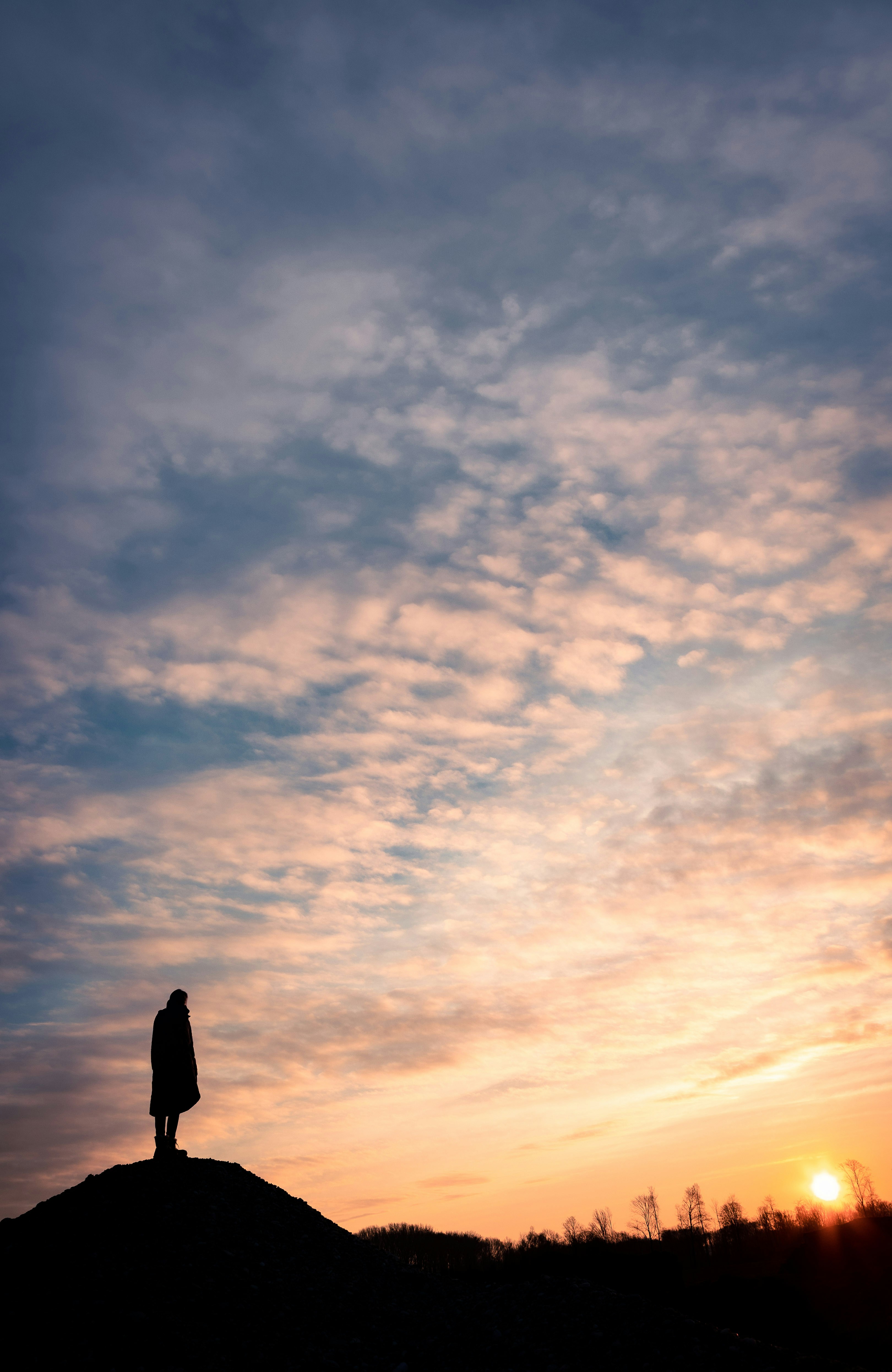A lone figure stands atop a hill, silhouetted against a vibrant sunrise and textured clouds. The scene conveys a sense of reflection and tranquility.