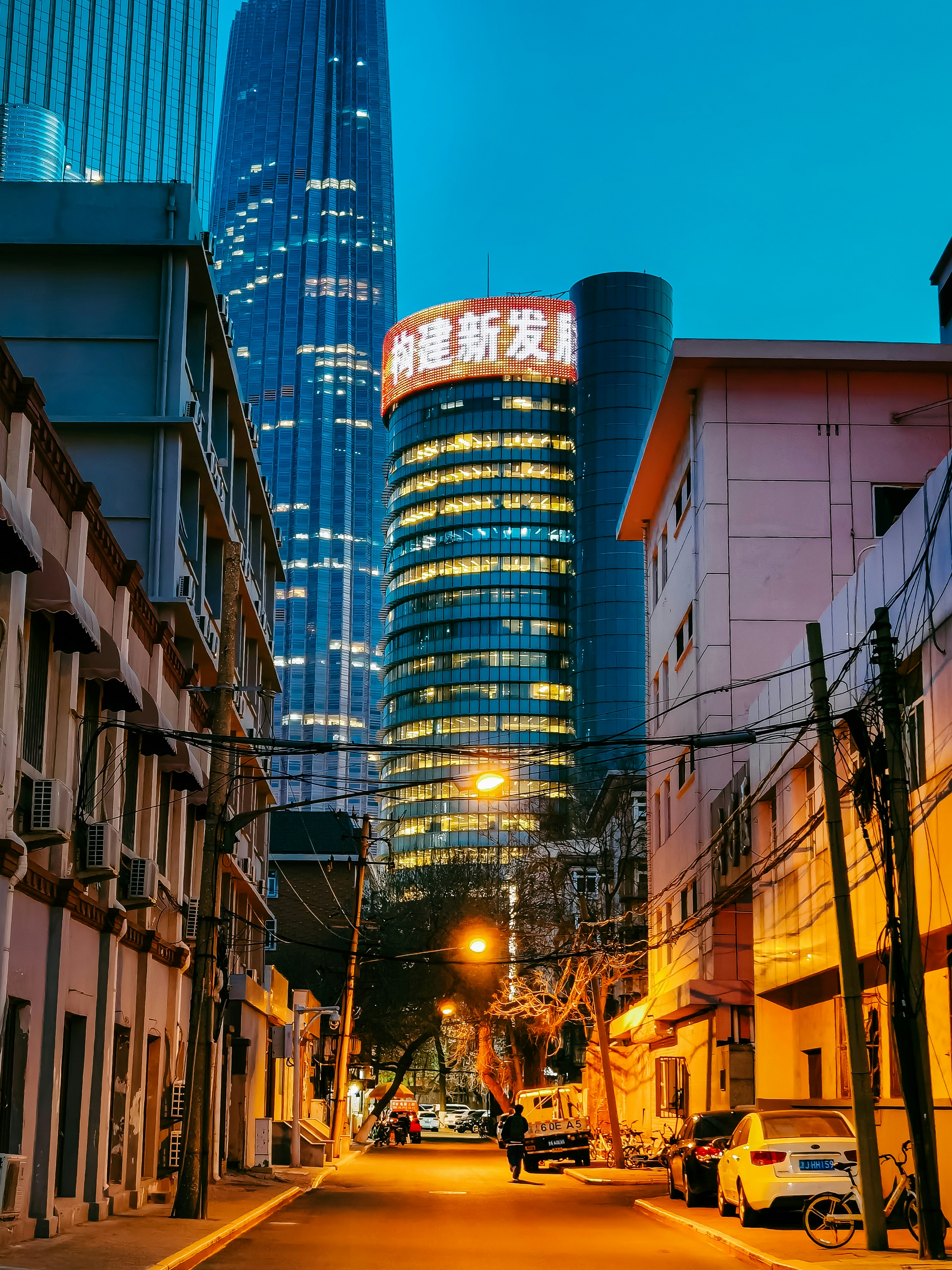 Illuminated street scene showcasing modern skyscrapers and vintage architecture under a twilight sky. Neon signage adds vibrant color to the urban landscape.