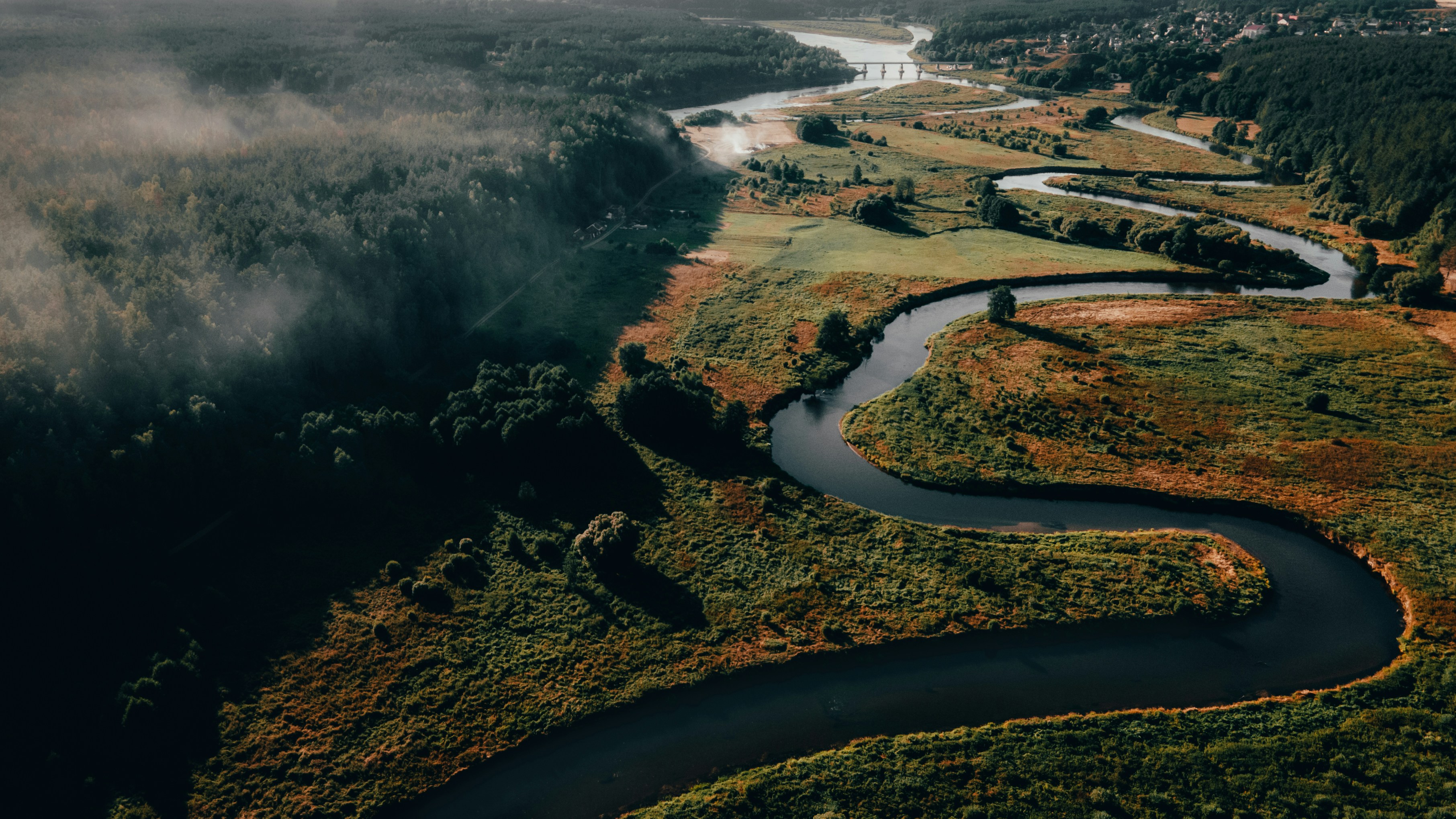 A winding river flows through lush green fields under a foggy sky, with distant hills lining the horizon.