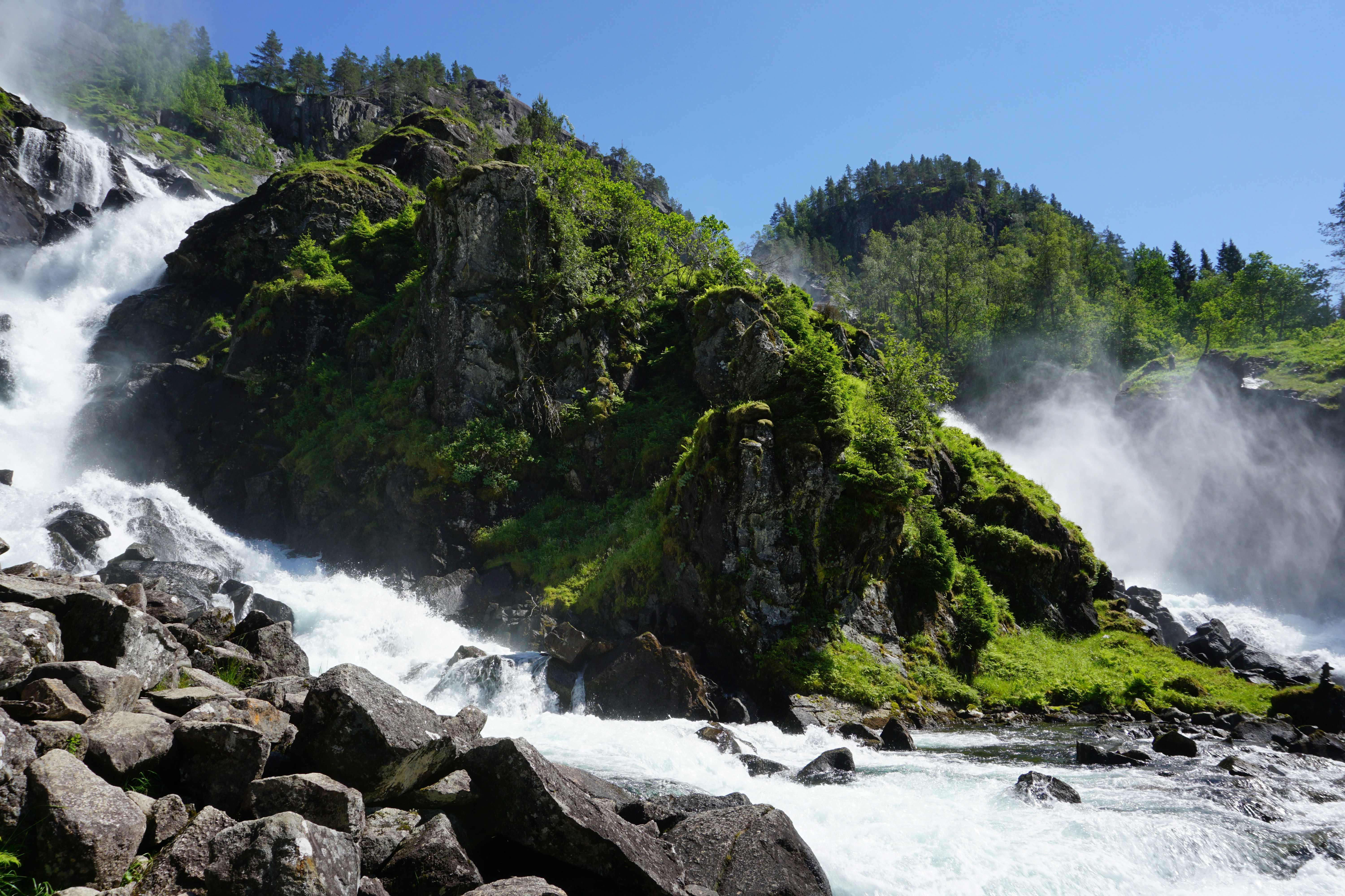 Rushing waterfall cascading over moss-covered rocks with lush greenery under a clear blue sky.