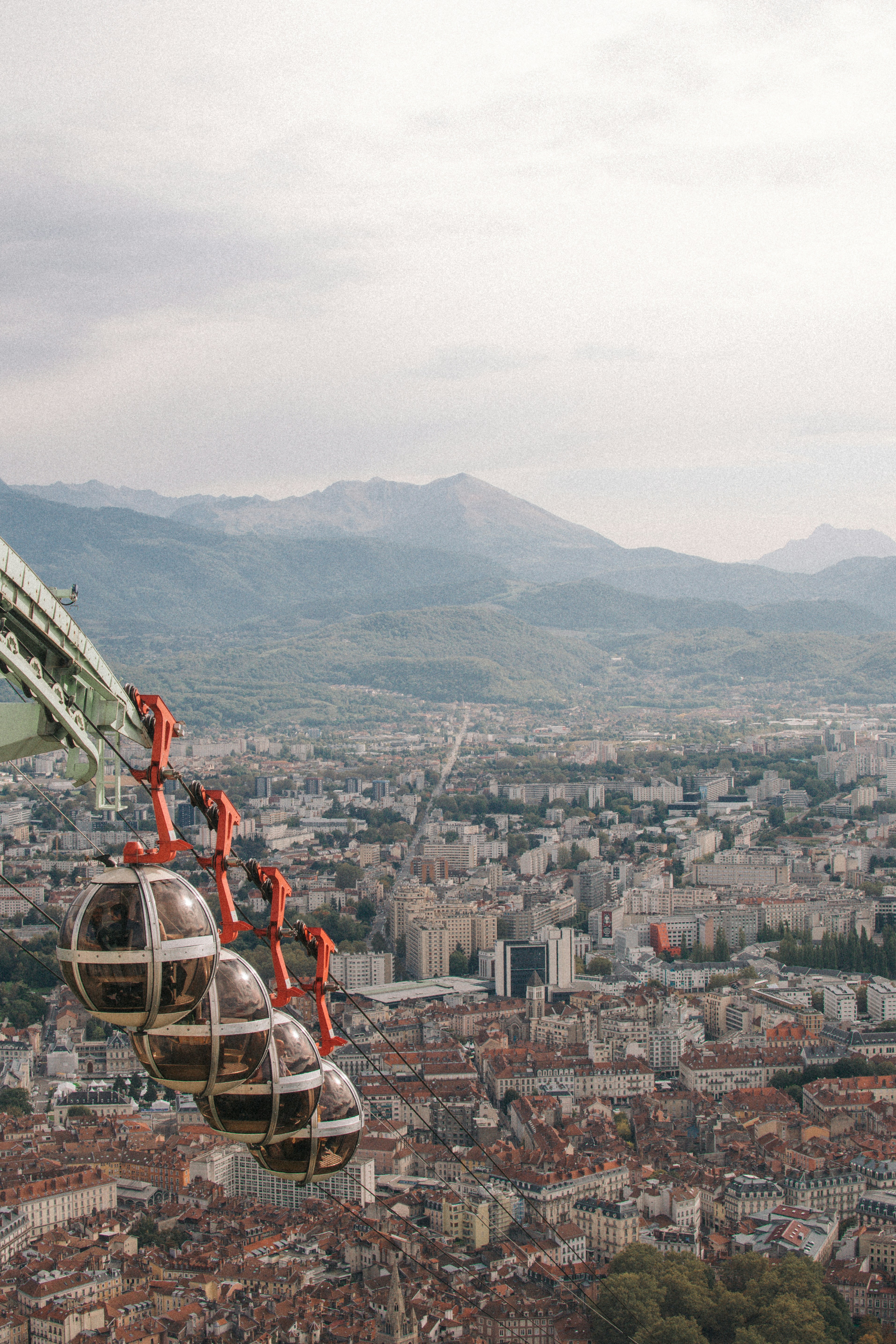 Cable cars glide over the cityscape of Grenoble, framed by distant mountains under a cloudy sky.