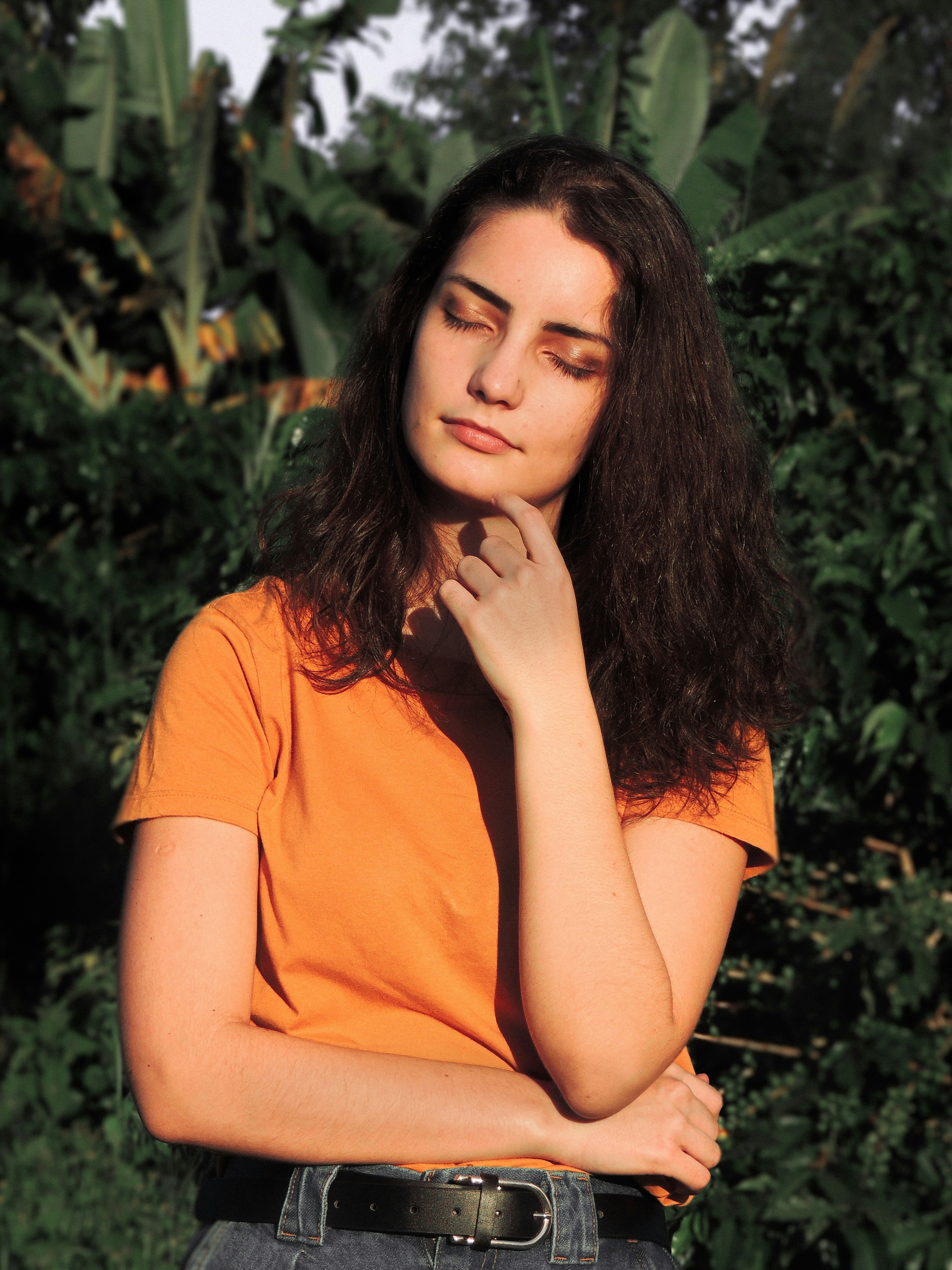 Woman in an orange shirt stands thoughtfully in a sunlit garden with lush greenery.
