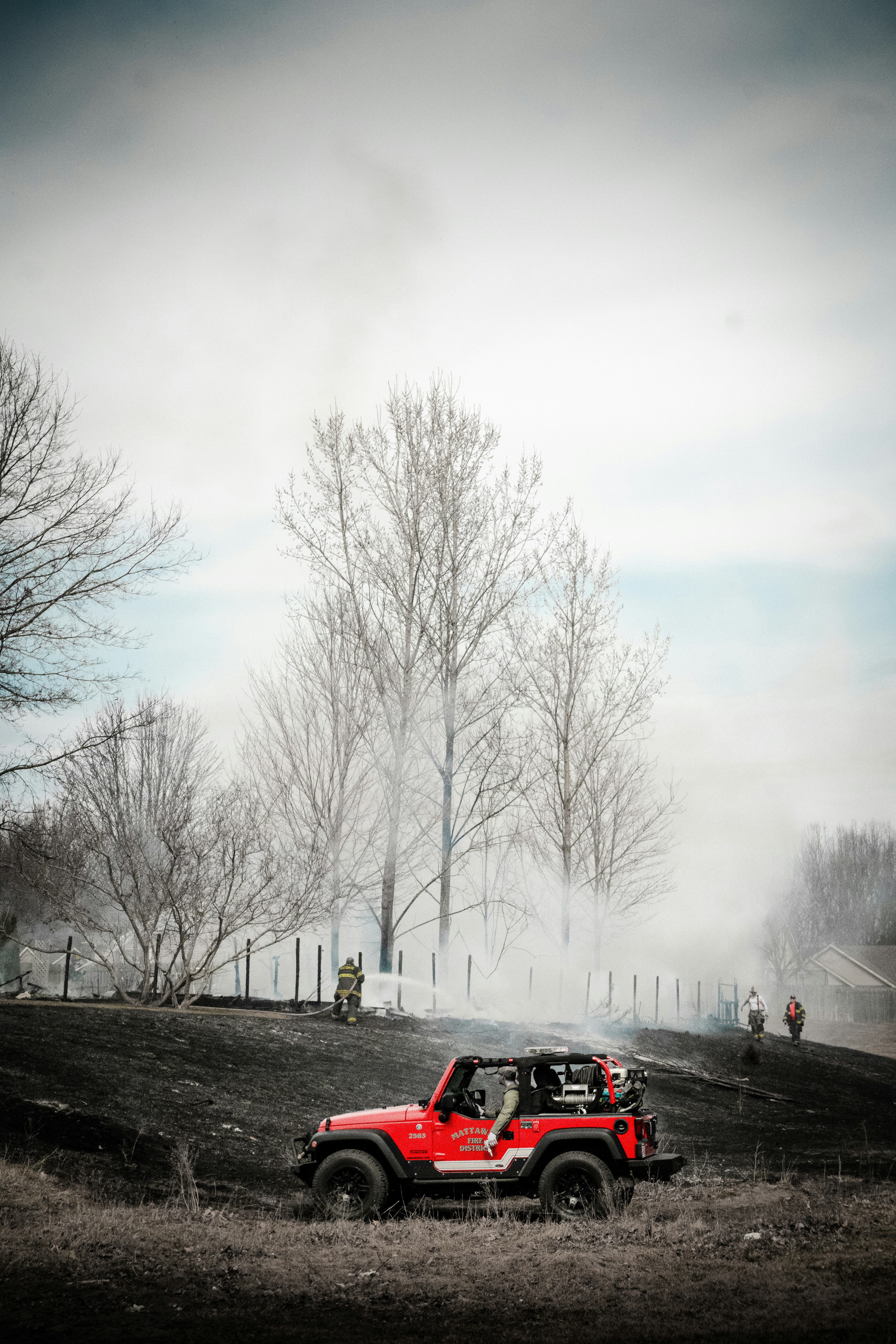A red Jeep stands amidst a charred landscape, with firefighters battling smoke in the background. The contrast between the vehicle and the scorched earth highlights the urgency of the situation.