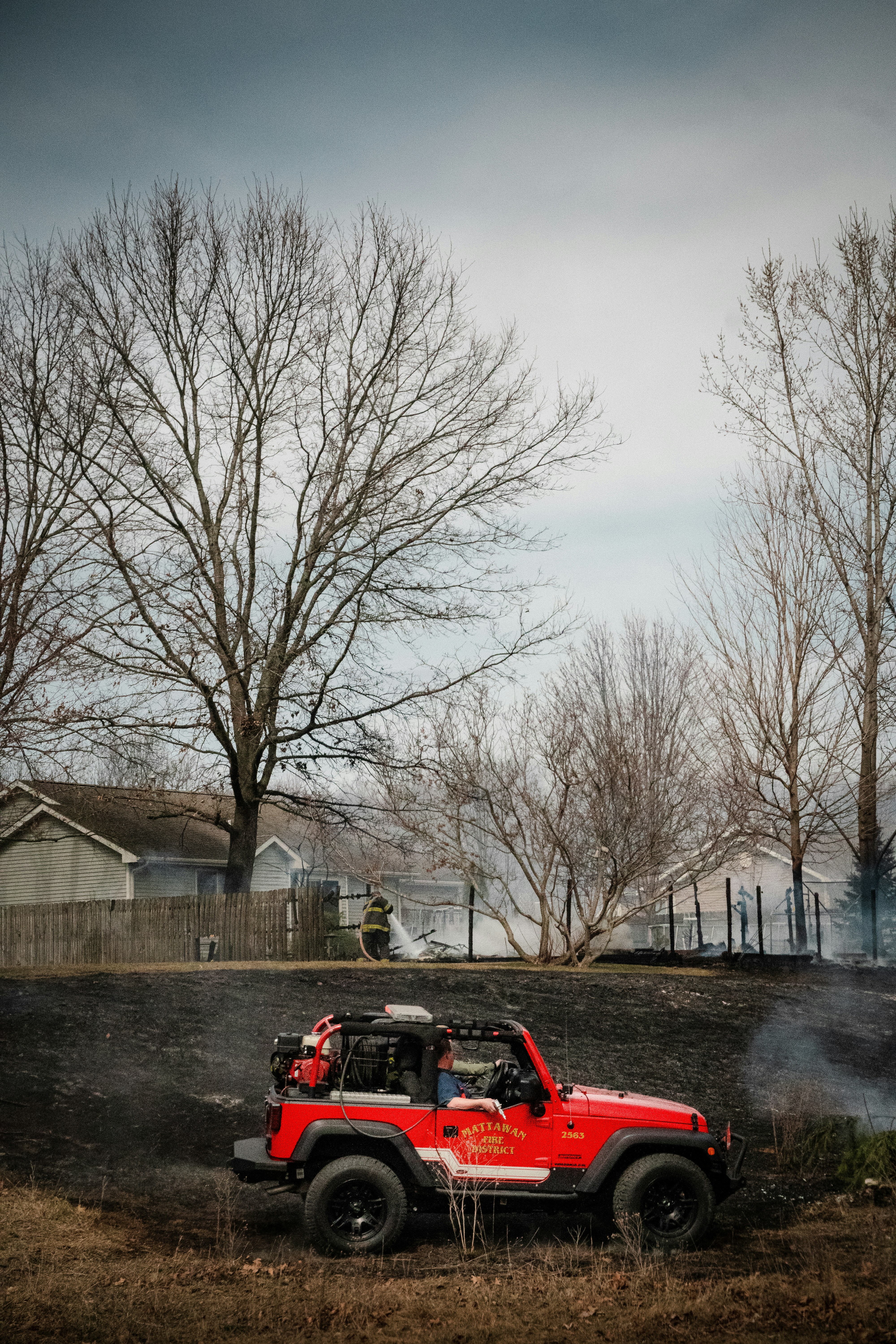 Firefighting vehicle navigating a scorched landscape, with trees and smoke in the background. A firefighter can be seen in action, emphasizing the urgency of the situation.