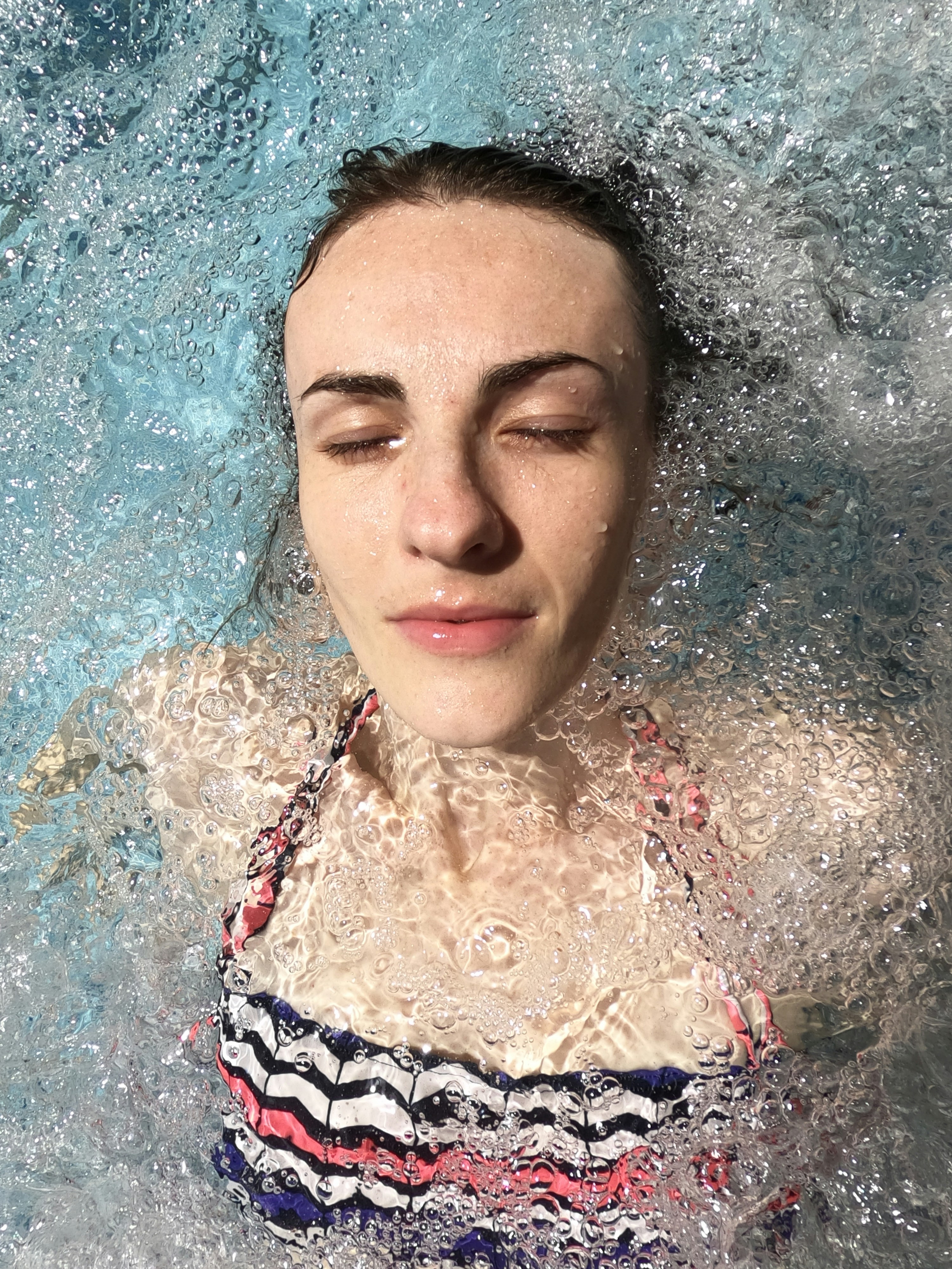 Close-up photograph of a woman with eyes closed, submerged in clear turquoise water as bubbles surround her, wearing a striped swimsuit.