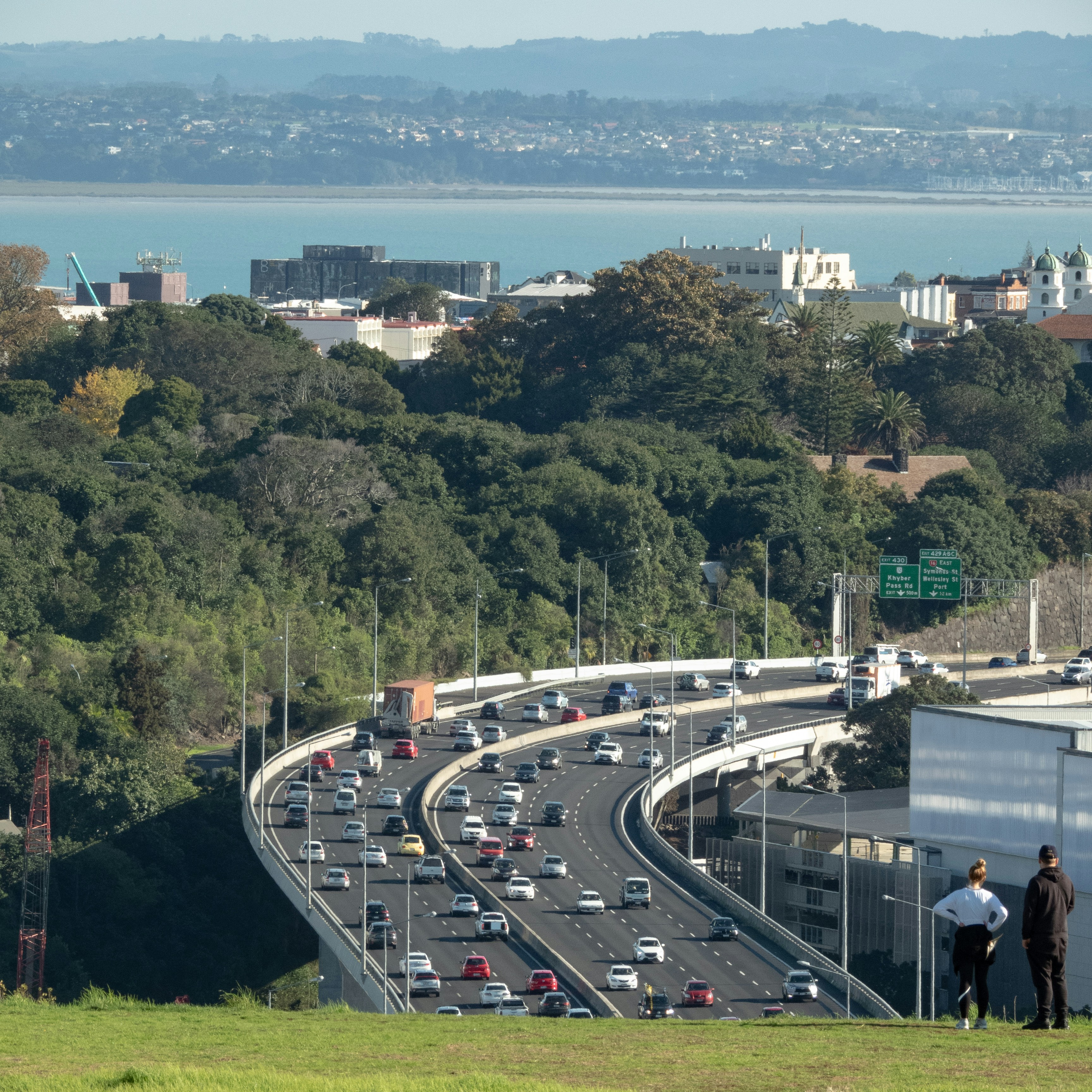 A busy highway curves through a lush landscape, with figures overlooking the scene. The contrast of urban infrastructure and natural greenery is striking.