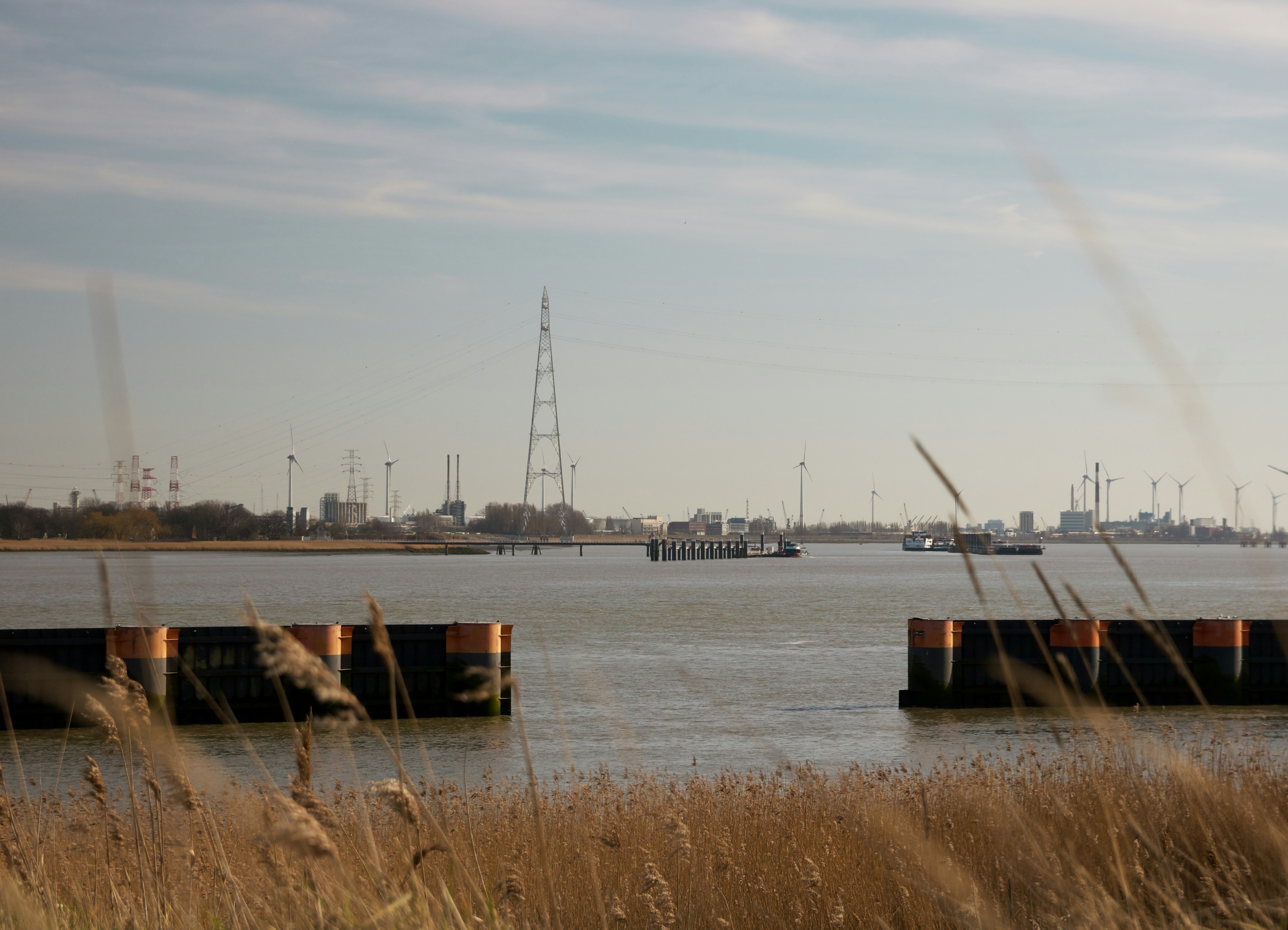 Grassy foreground frames a calm river with industrial structures and a city skyline in the distance.