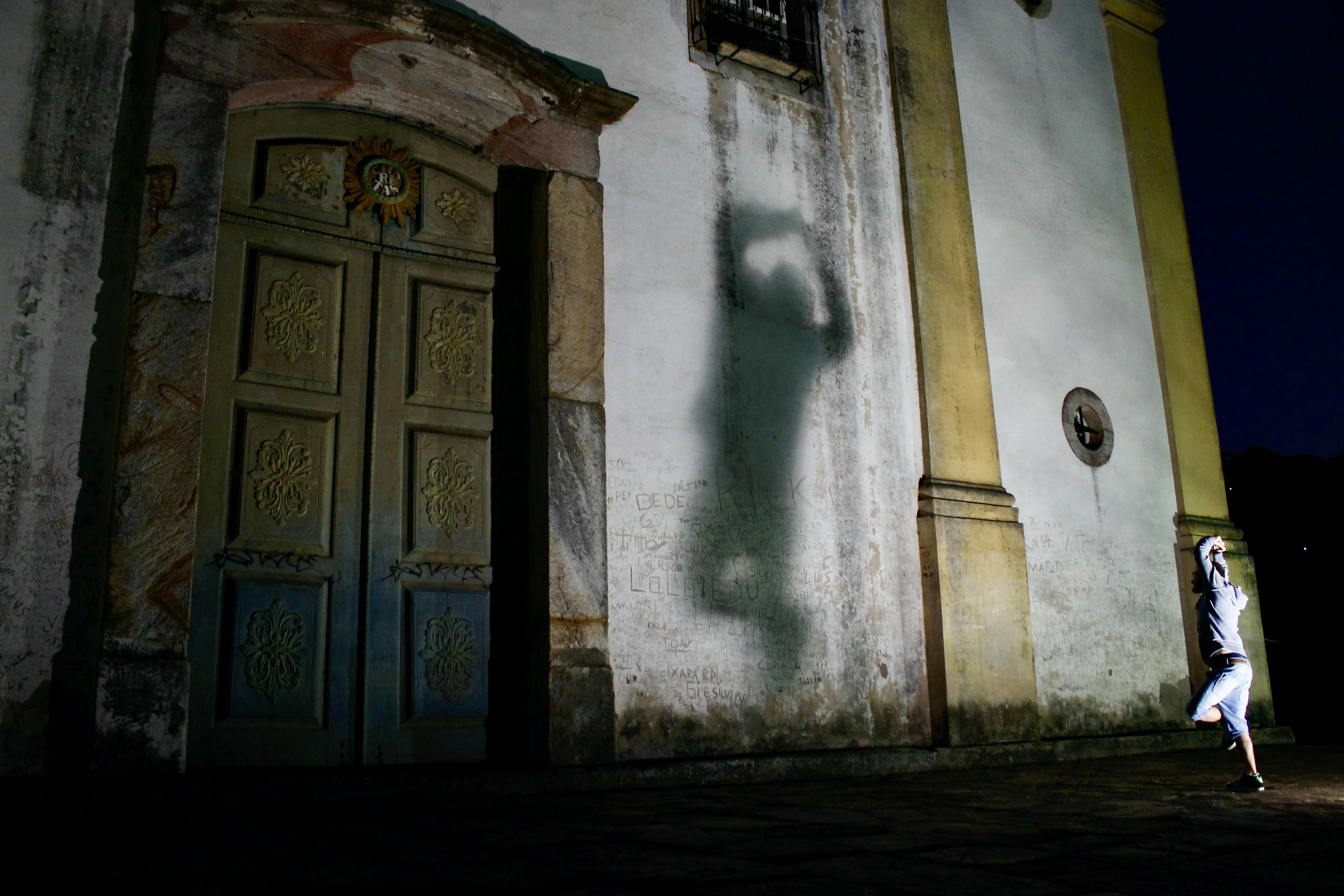 A person in white walks beside a historic building with a shadow cast against the wall.
