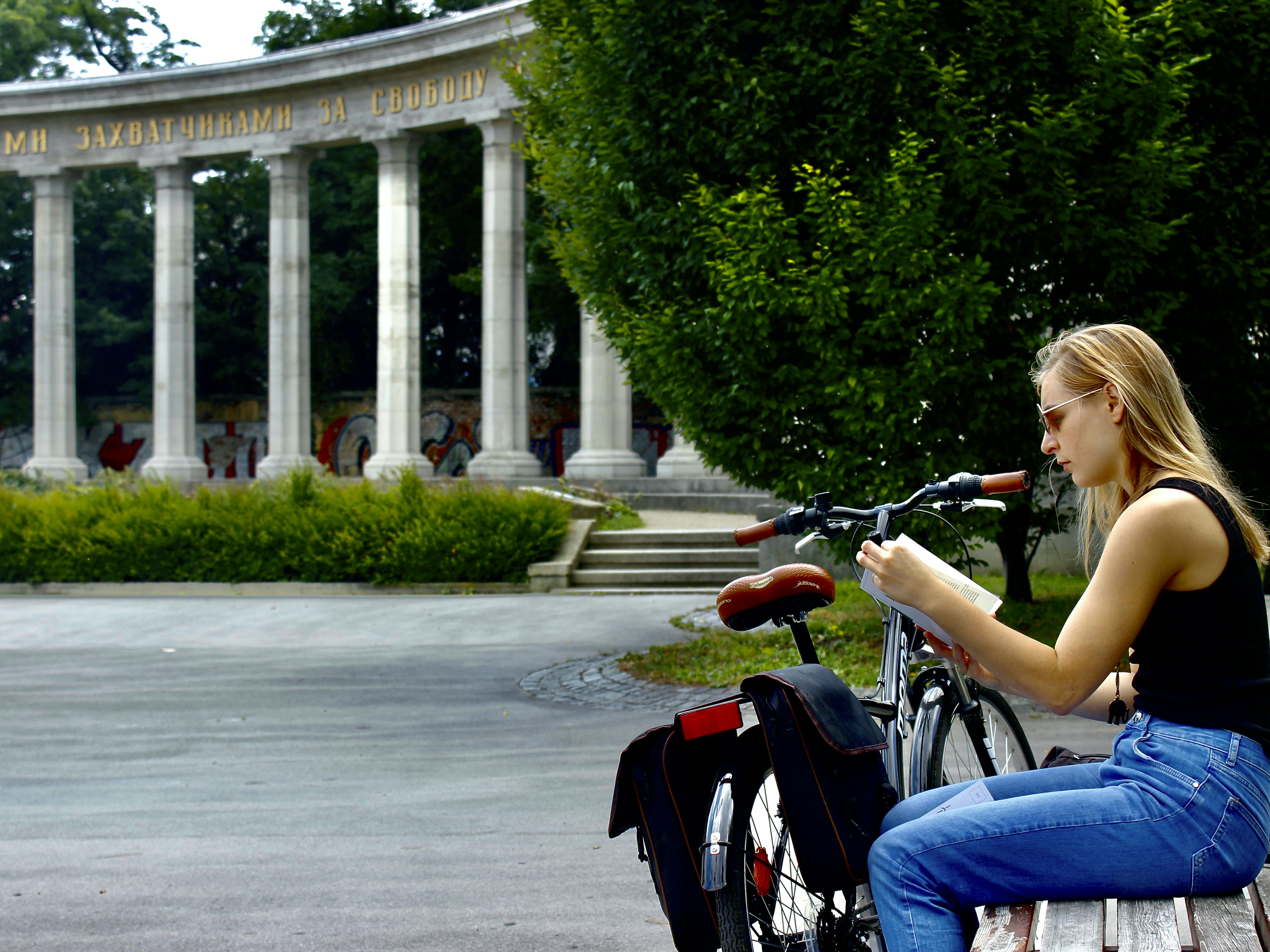 A young woman sits on a bench, engrossed in her phone, with bicycles nearby and a classical colonnade in the background. The scene captures the blend of nature and urban architecture.