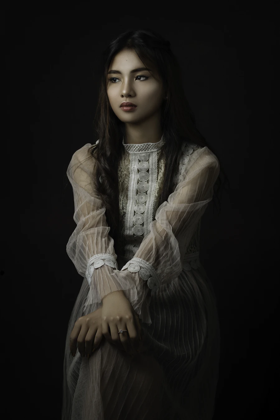 A woman seated against a black background, wearing a delicate sheer dress with lace detailing and looking off to the side. Her hands rest in her lap, with soft studio lighting shaping her face and clothing.