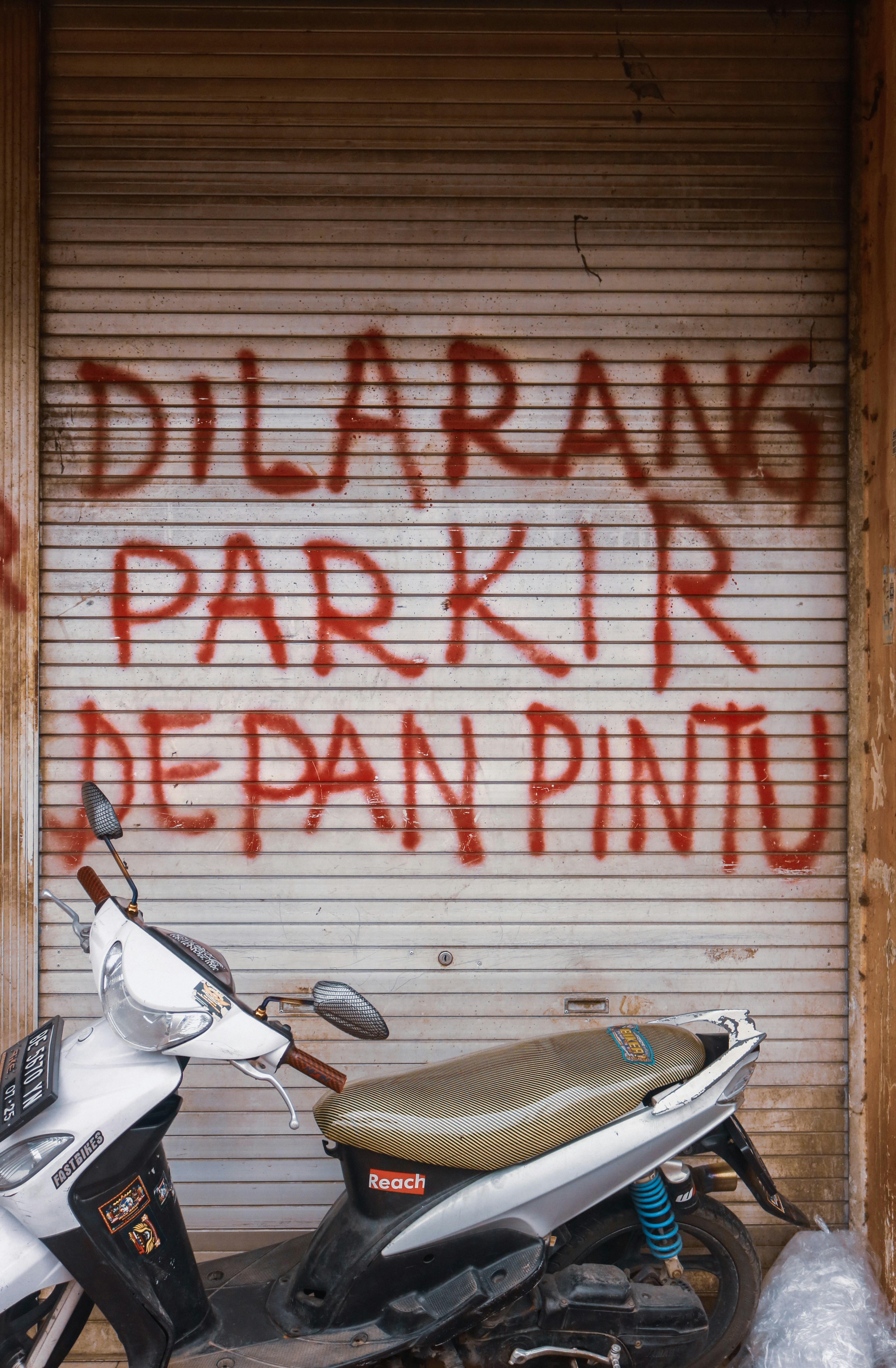 Graffiti stating 'DILARANG PARKIR DEPAN PINTU' on a corrugated metal shutter, accompanied by a parked scooter, reflecting urban culture and local regulations.