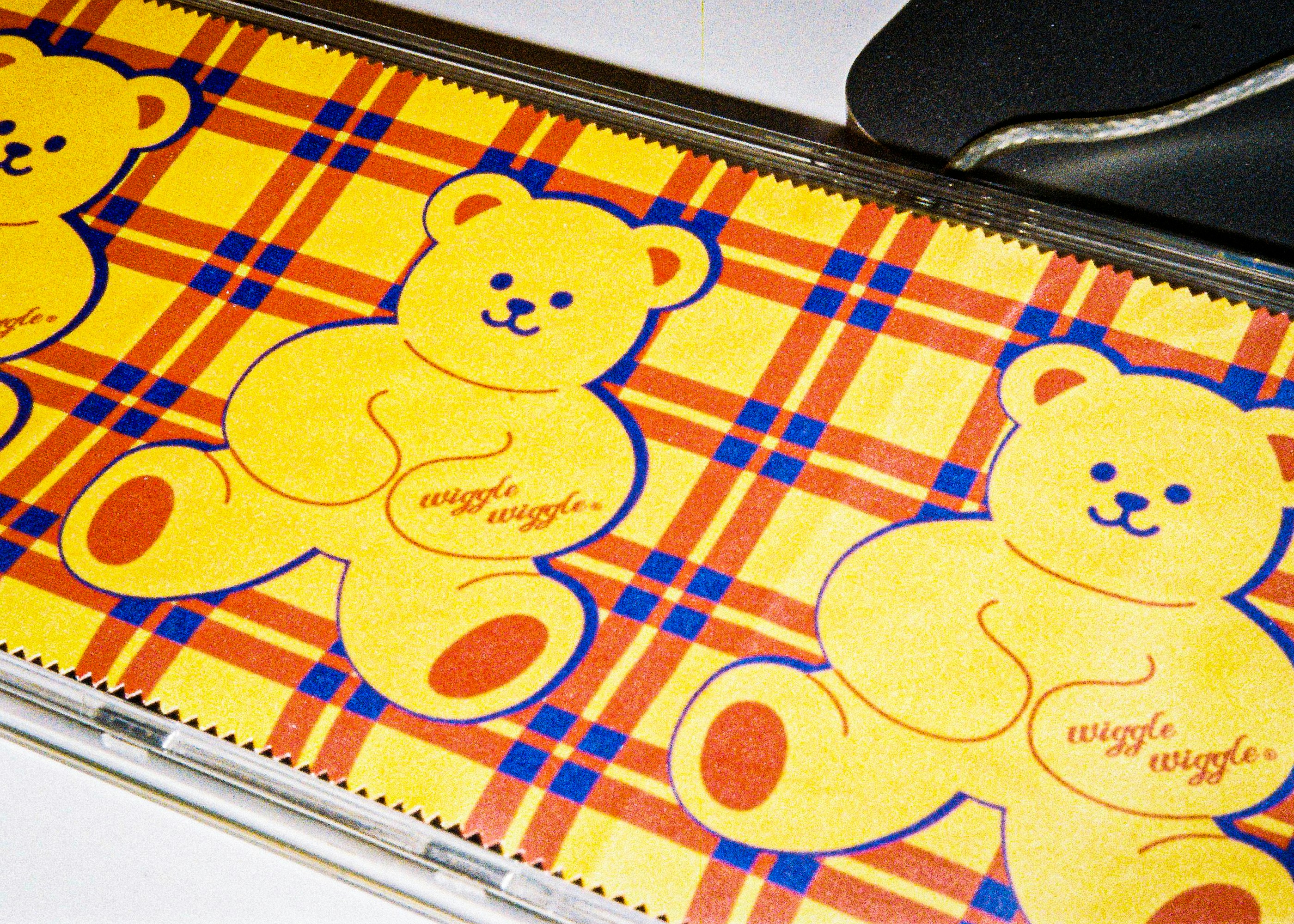 Close-up of cartoon teddy bears repeated on yellow decorative tape with an orange plaid background. The design is a printed illustration on clear packaging tape.