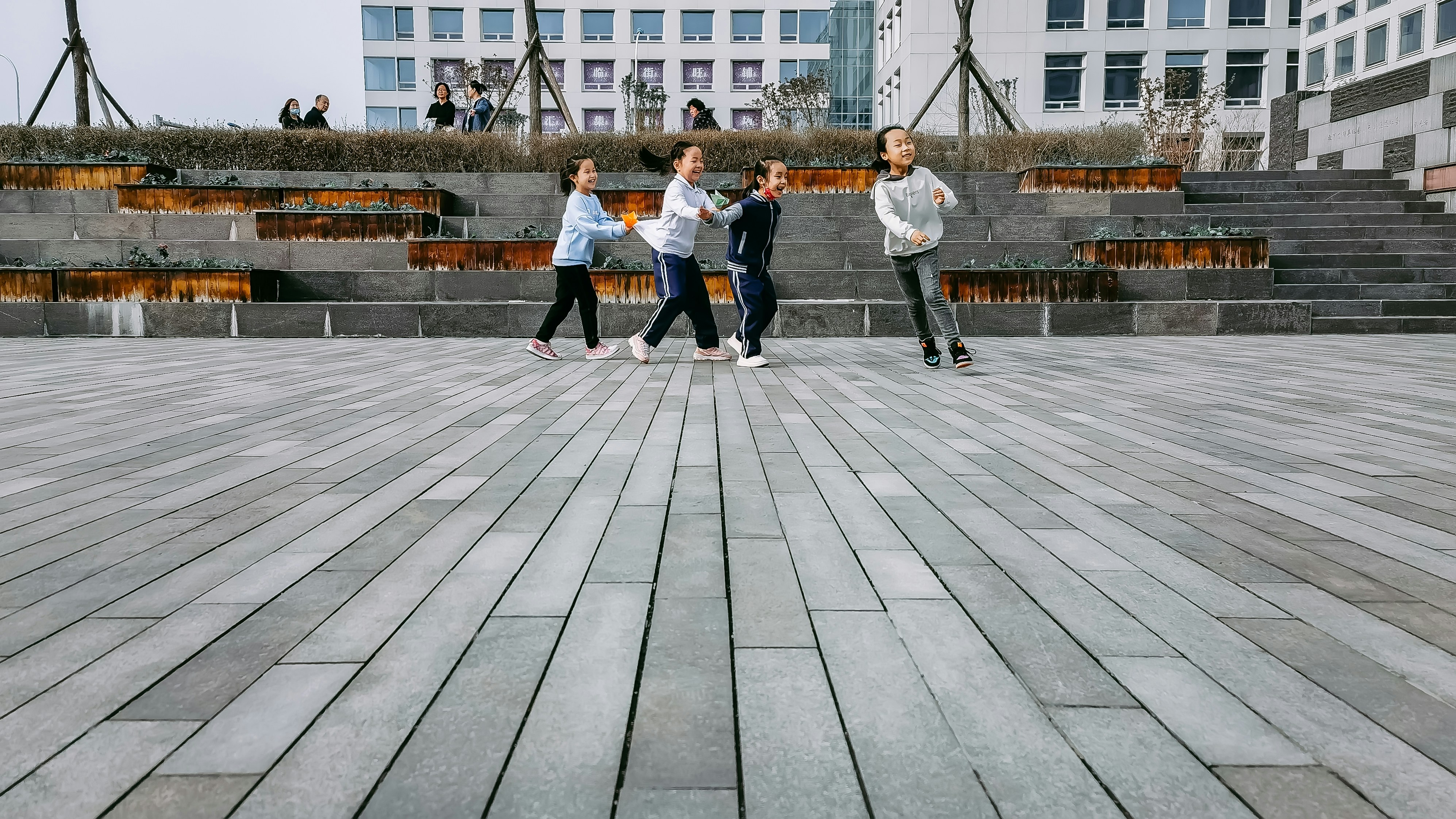 Four children engaging in playful activity on a modern stone plaza surrounded by urban architecture.