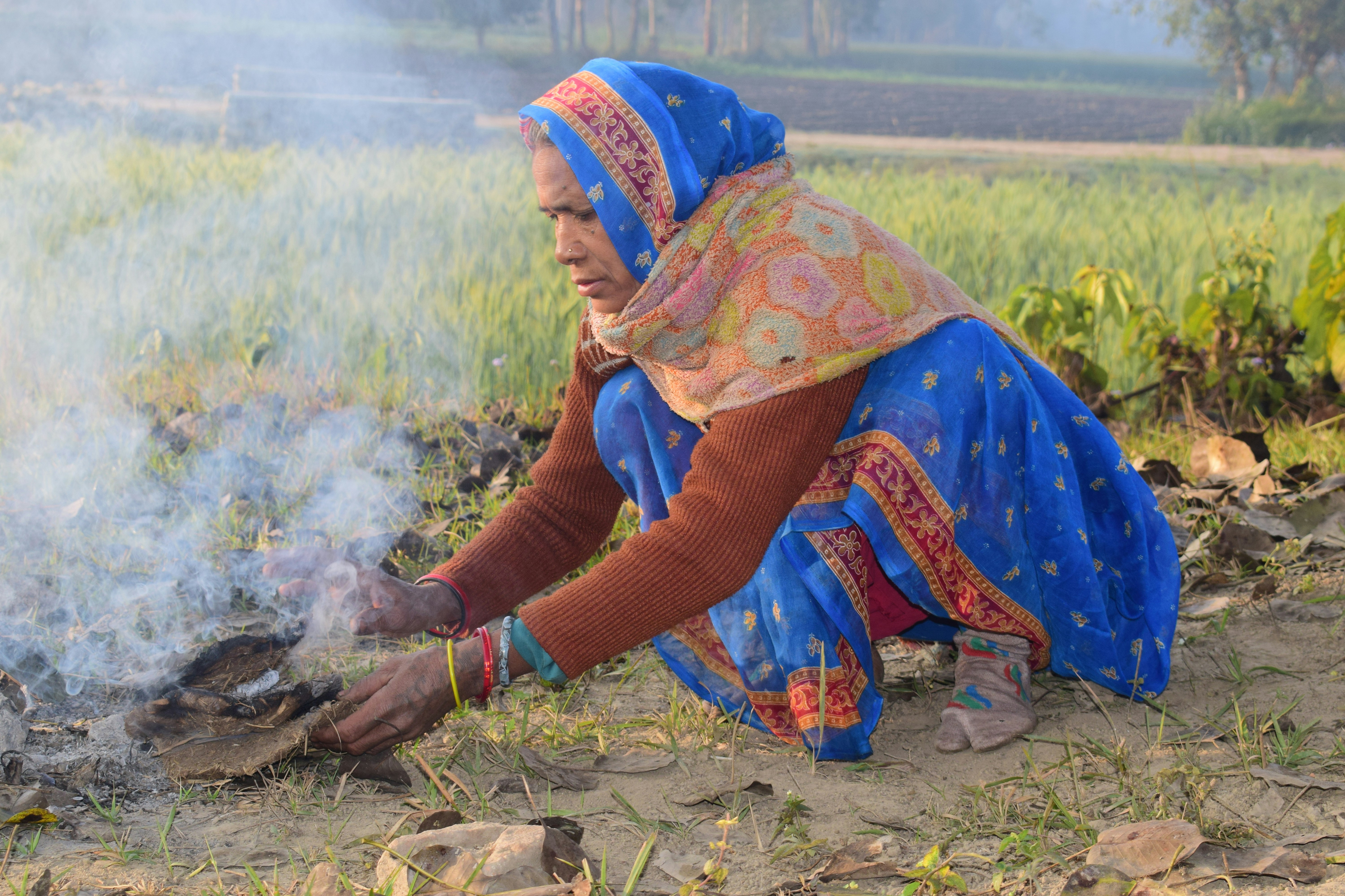 Woman in colorful attire tending a small fire amidst lush green fields with a hint of morning mist.