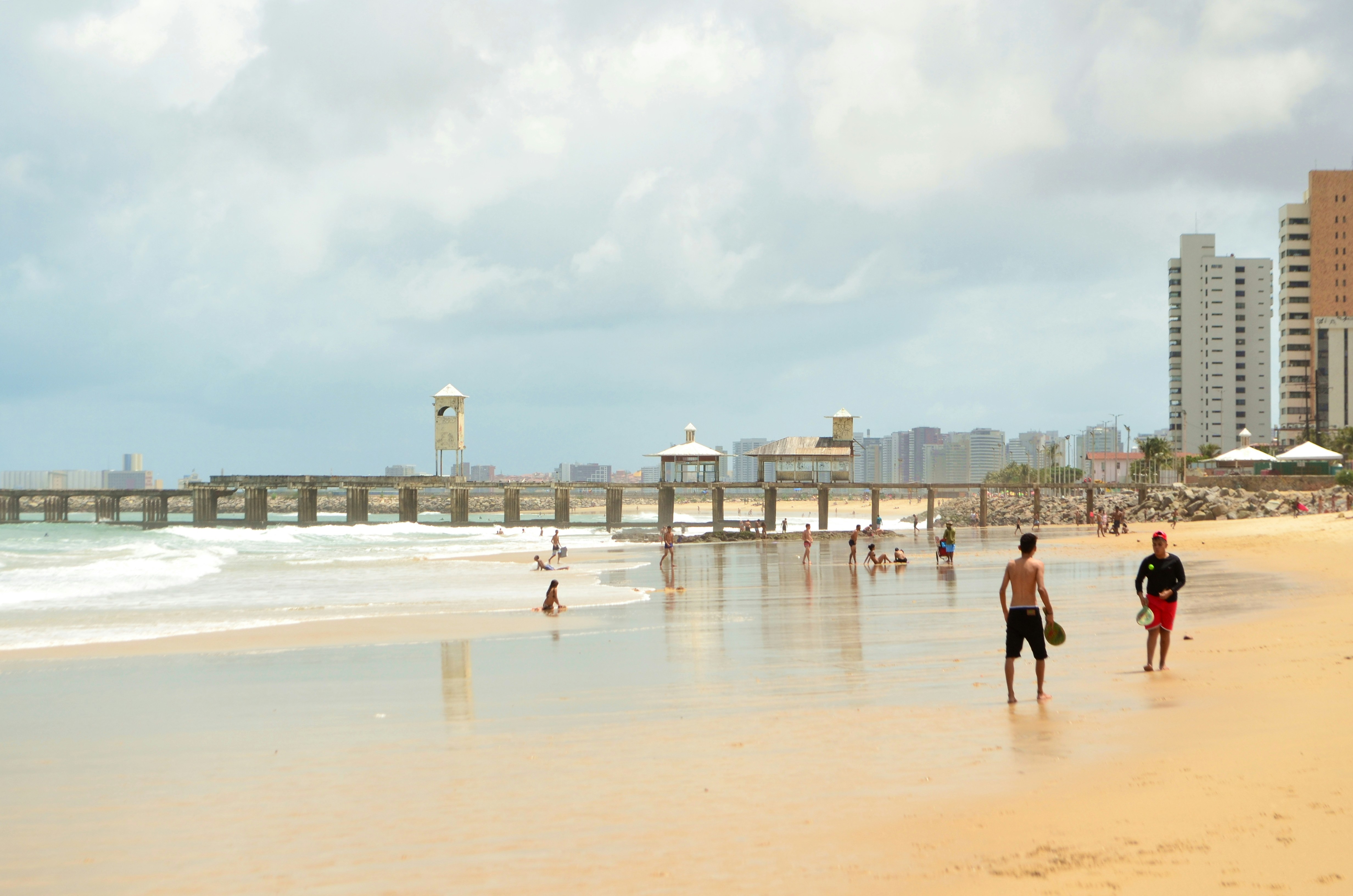 People walking along a sandy beach with waves and a distant pier under a cloudy sky.