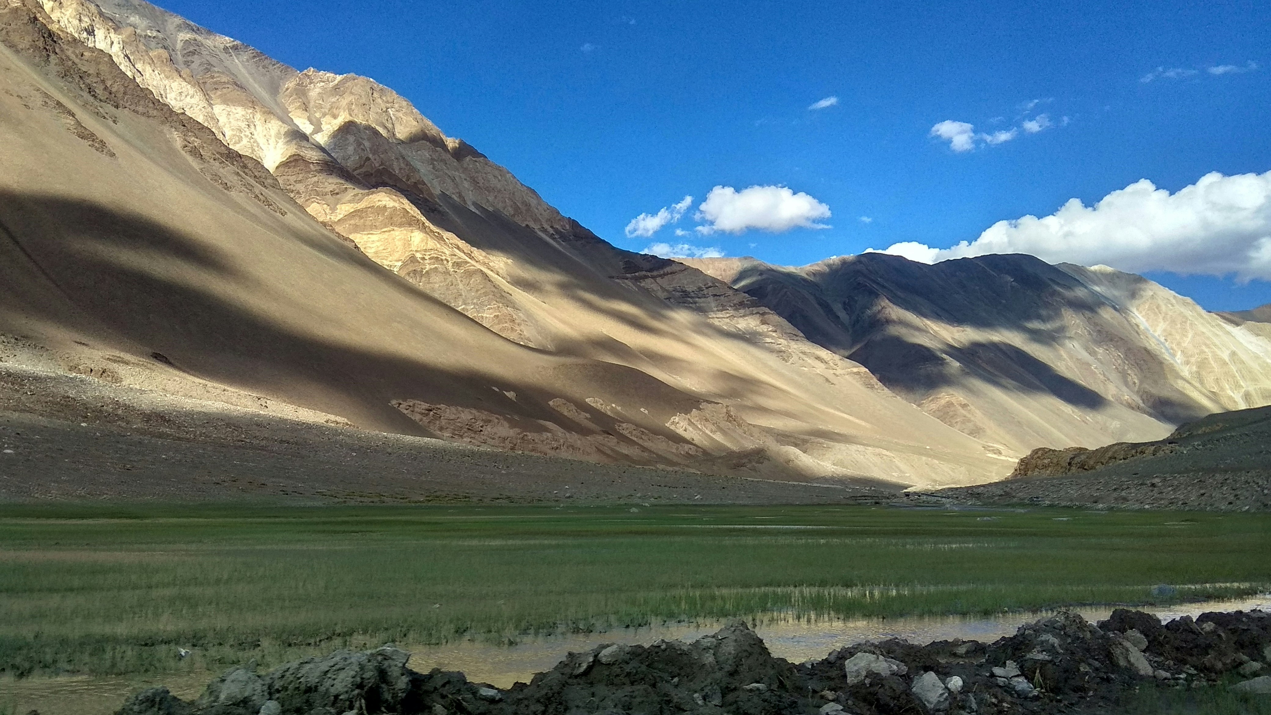 Vast mountain landscape with layered terrain and vibrant green fields under a clear blue sky. The shadows cast by the peaks create a dynamic interplay of light and color.