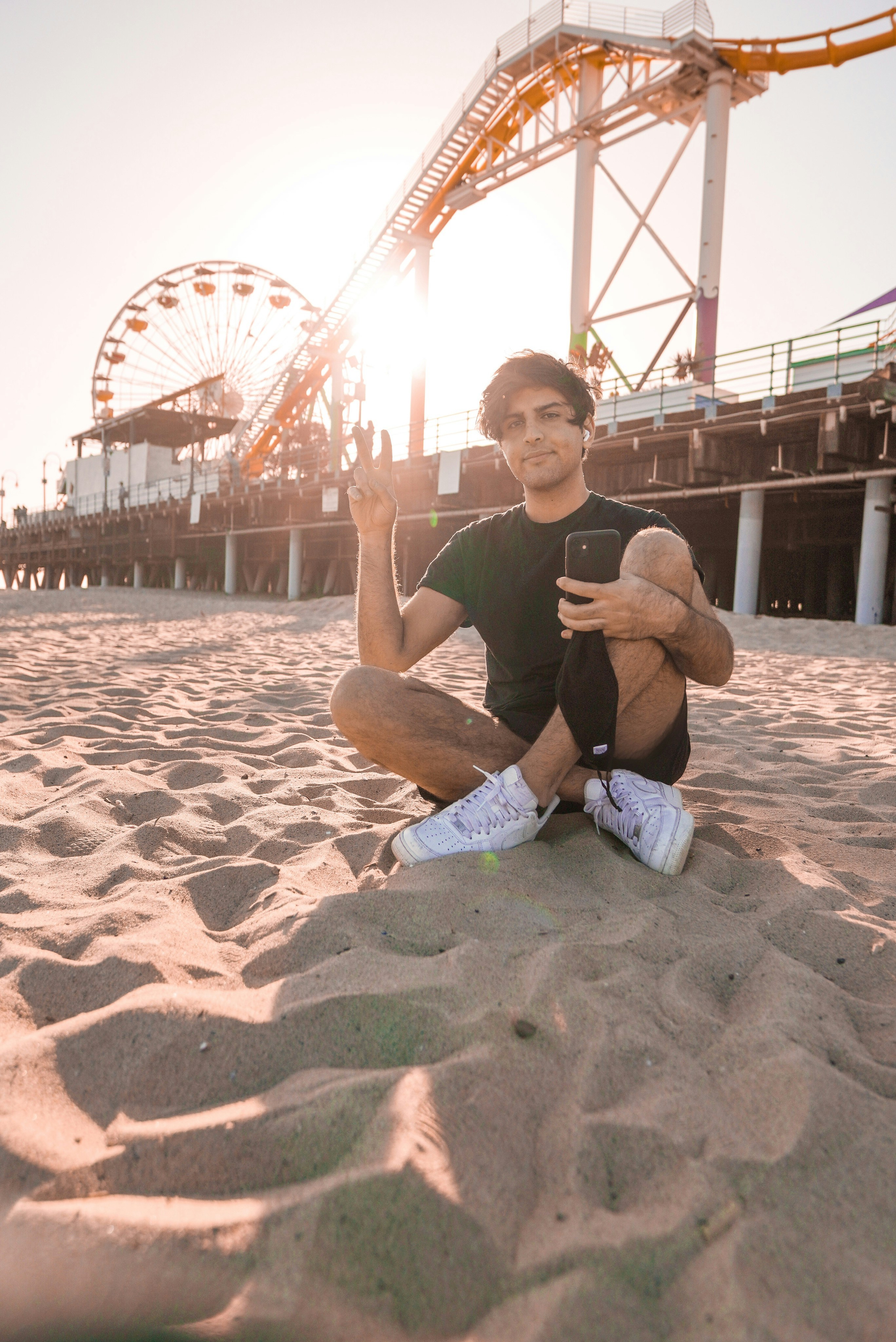 Sunlit Beach Portrait by the Pier: Man Seated on Sand with Ferris Wheel and Roller Coaster Backdrop
