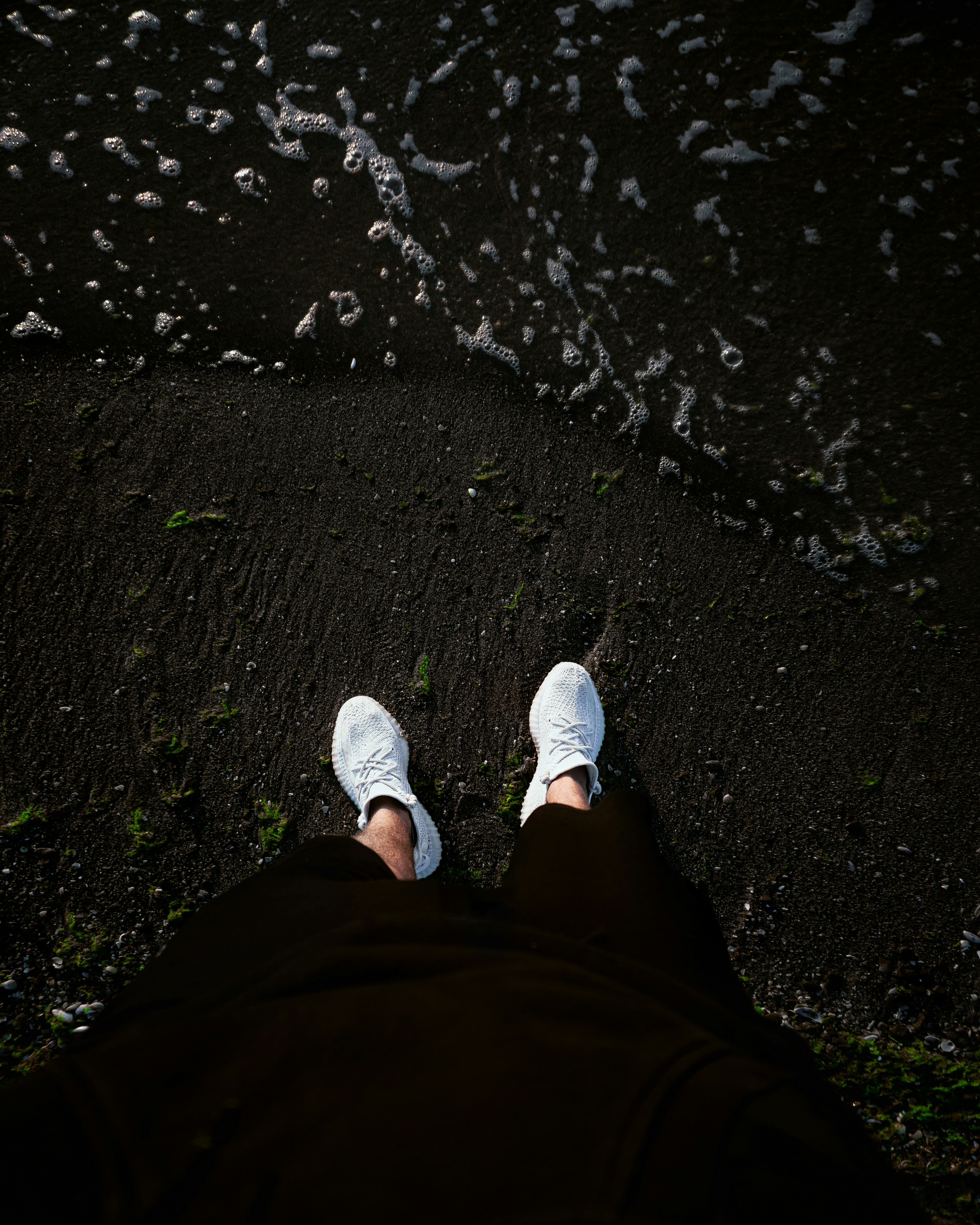 Aerial view of a person standing on the shore, with white sneakers partially submerged in ocean waves on a dark sandy beach.